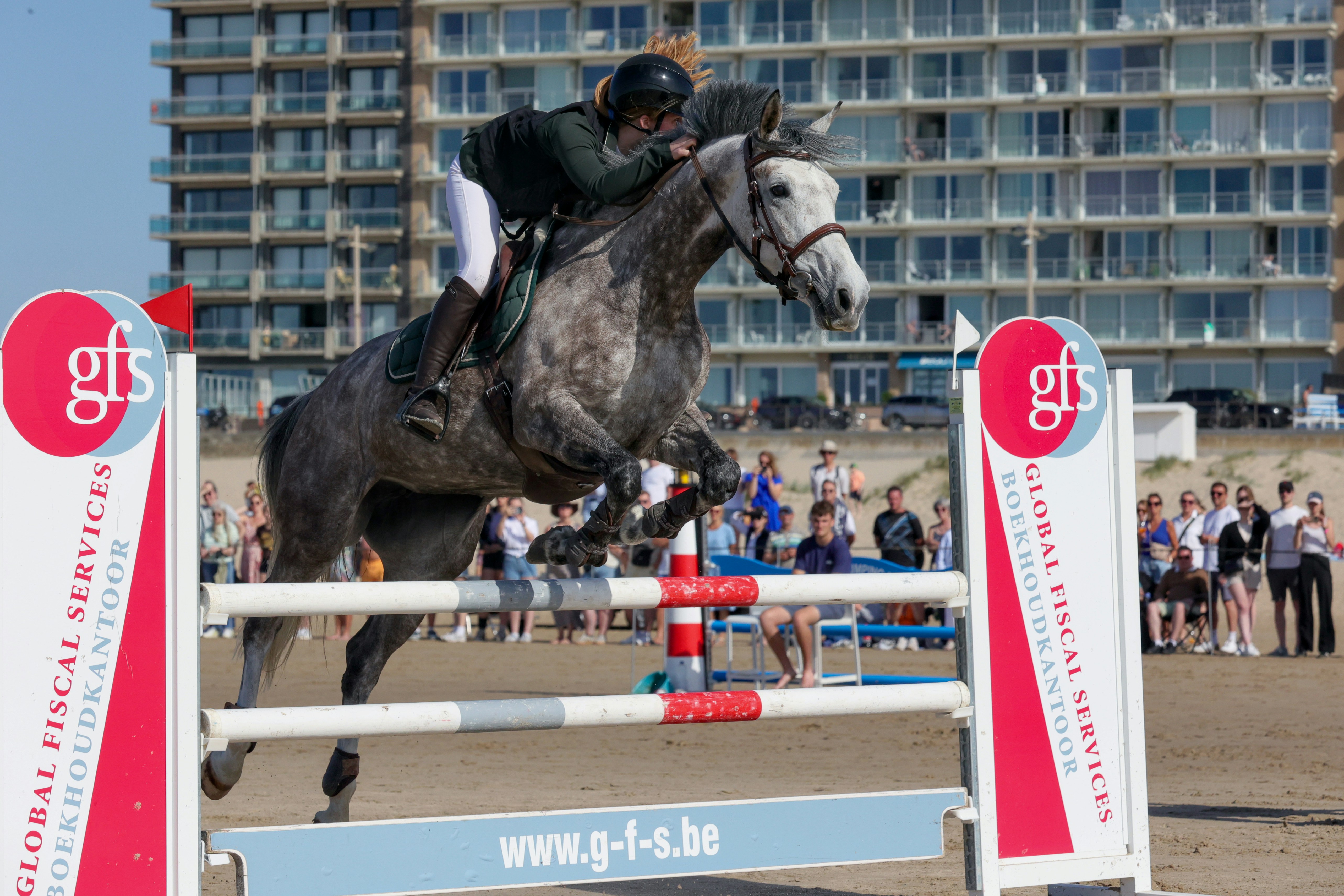 Rider and Grey Steed Clear a Jump Against a Crowded Beachside Arena