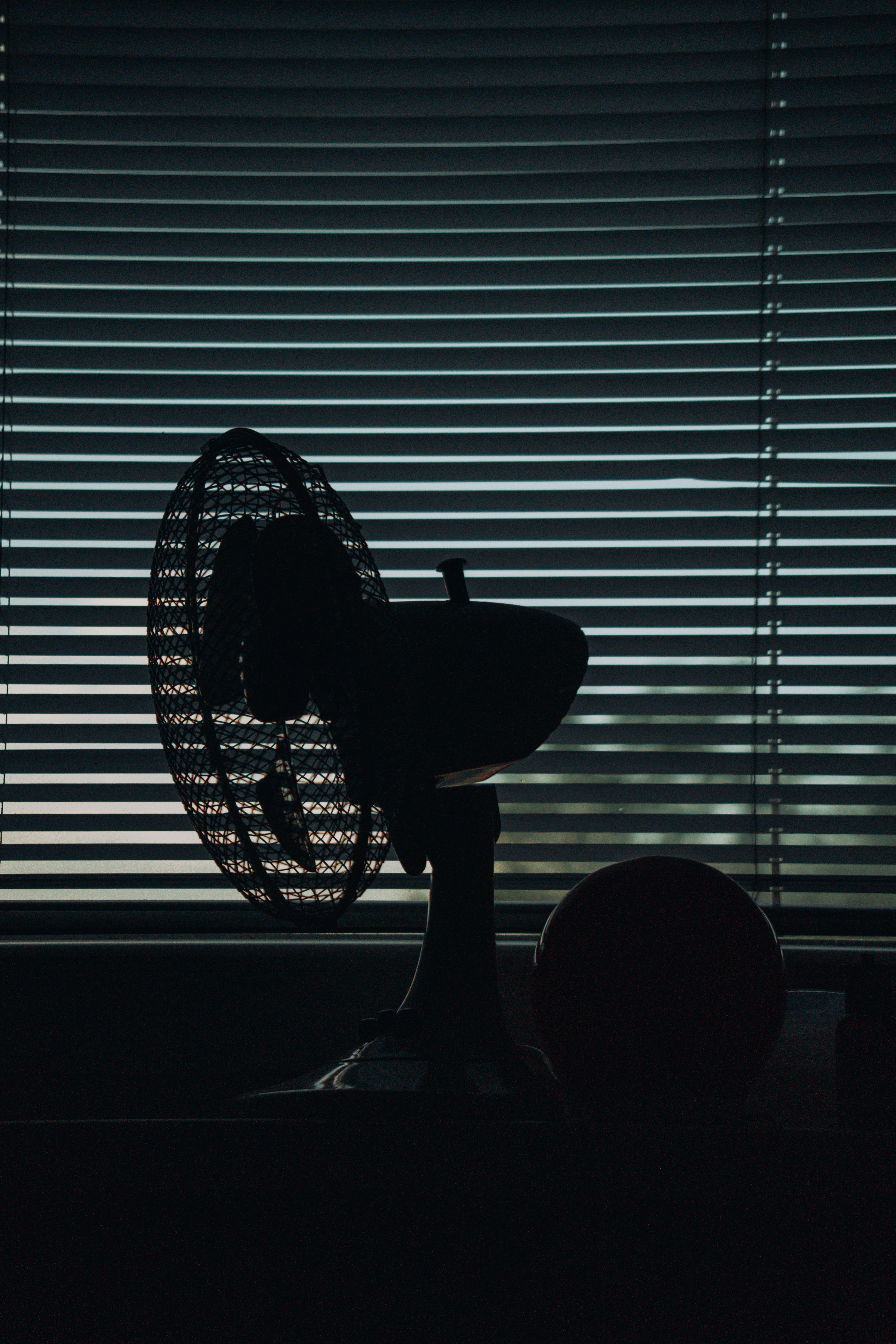 A fan and object silhouetted against window blinds.