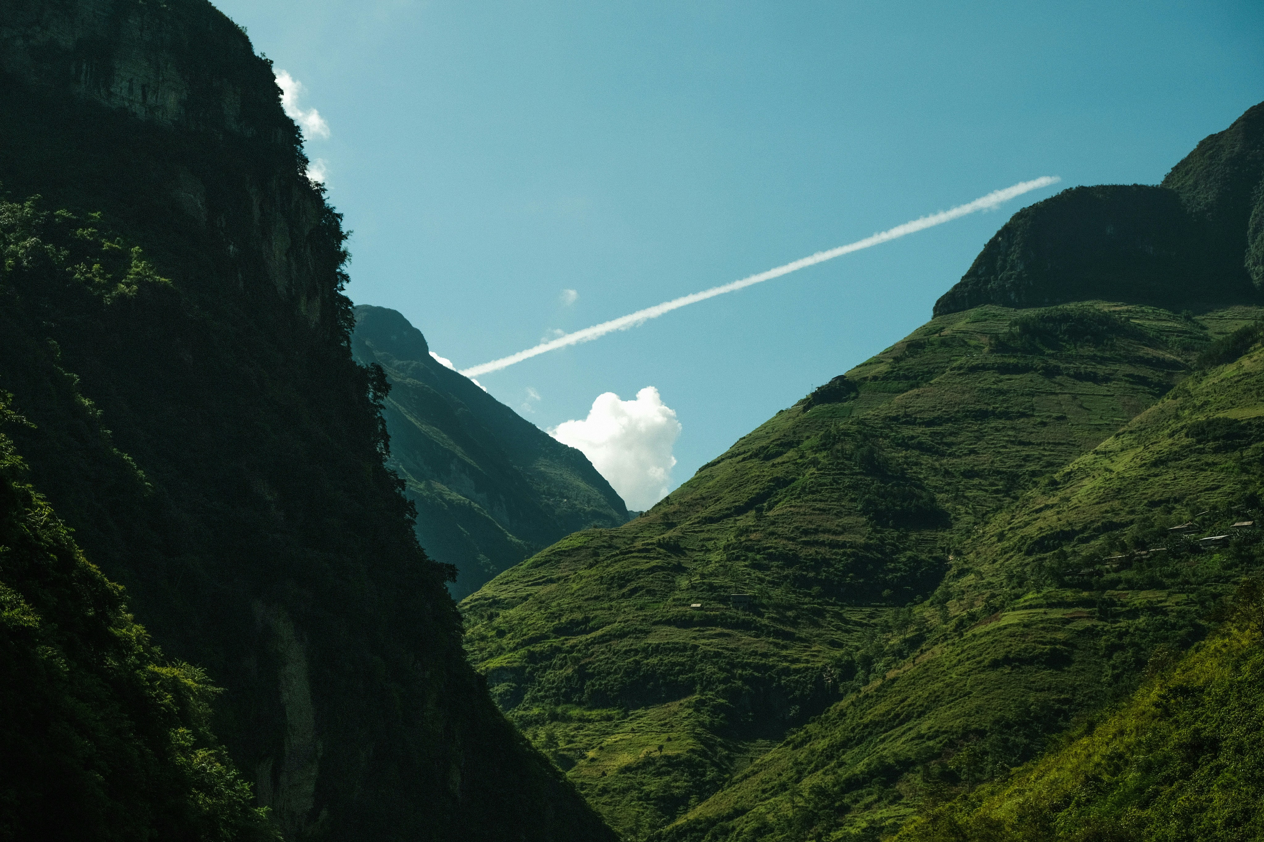 Sunlit green mountains frame a deep valley with a bright contrail crossing a blue sky, emphasizing scale and depth.