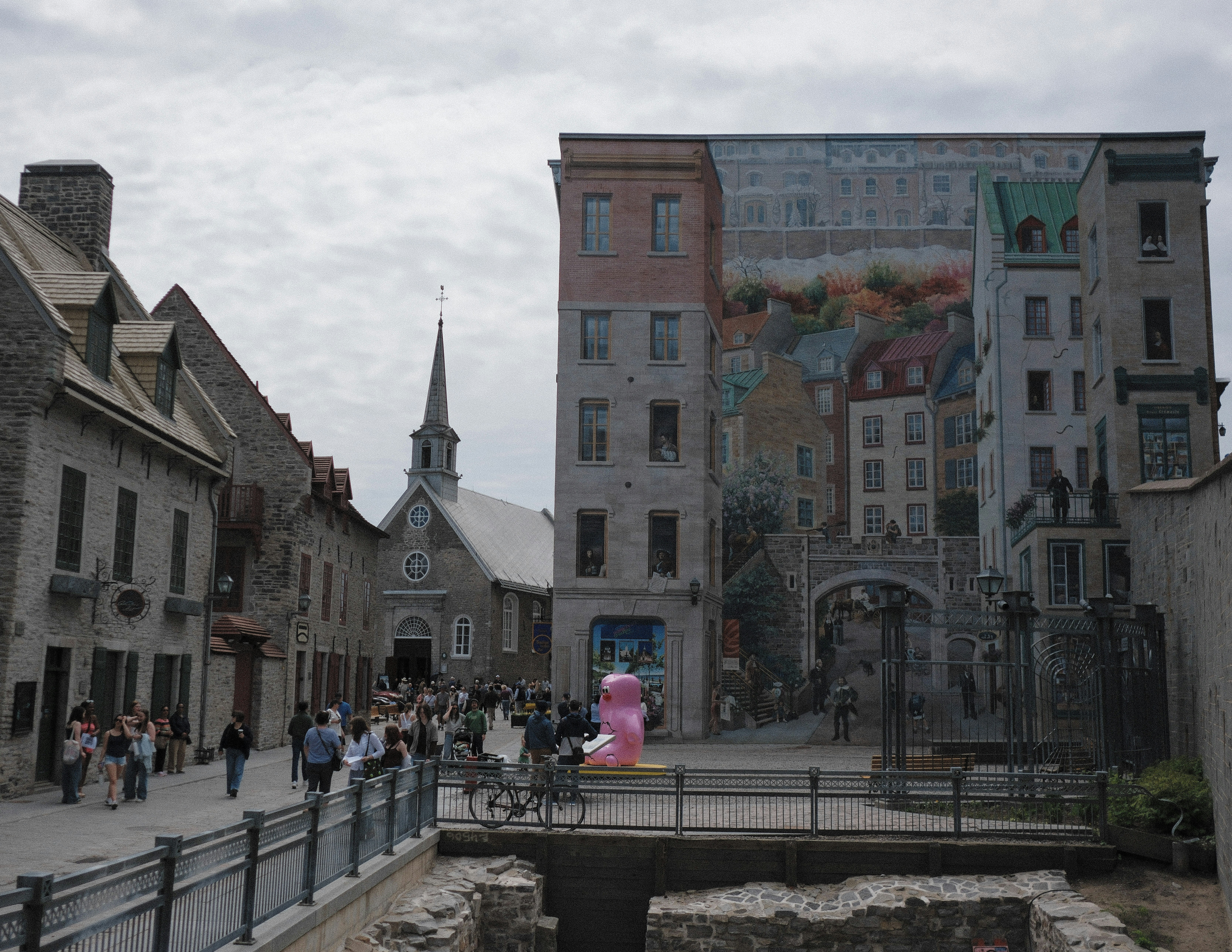 A high-angle photograph captures a bustling public square featuring a massive, detailed mural painted on the side of a tall building. The artistic mural blends seamlessly with real architecture, showing historic scenes and colorful fall trees. In the square below, crowds of people gather near a small church with a pointed spire, while a playful pink sculpture adds a pop of modern color. This photo was taken by Fujifilm xt5 with 23mm and no edit just fuji color, showcasing a perfect blend of historic community life and urban street art.
