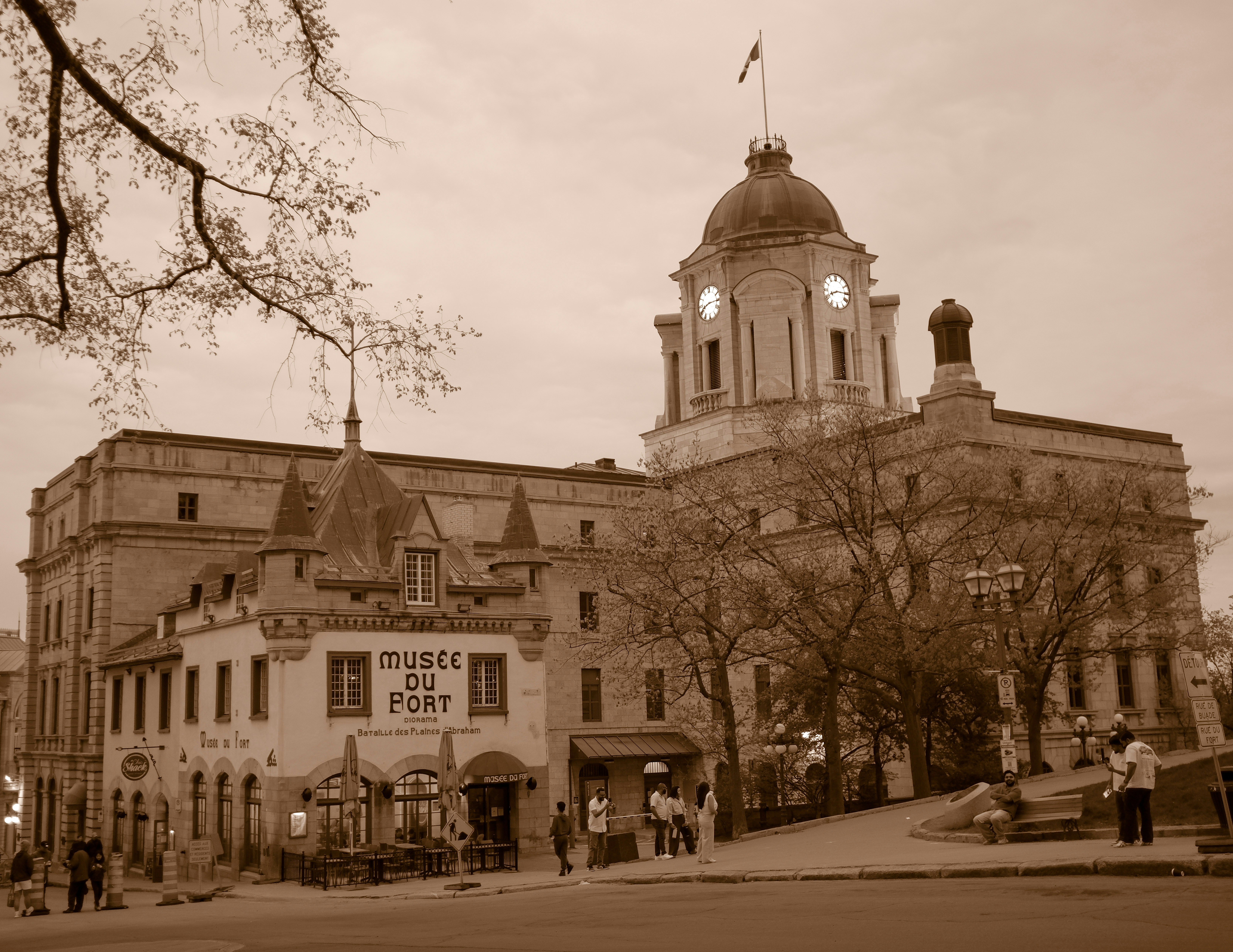 An artistic sepia-toned photograph captures the historic Musée du Fort building on a city street corner. The charming building features chateau-style towers, while a grand government structure with a prominent clock tower and a flag waves in the background. A few people relax on benches along a sloping sidewalk under bare tree branches. This photo was taken by Fujifilm xt5 with 23mm and no edit just fuji color, using a classic warm tone to emphasize the vintage texture and timeless architectural art of the city.
