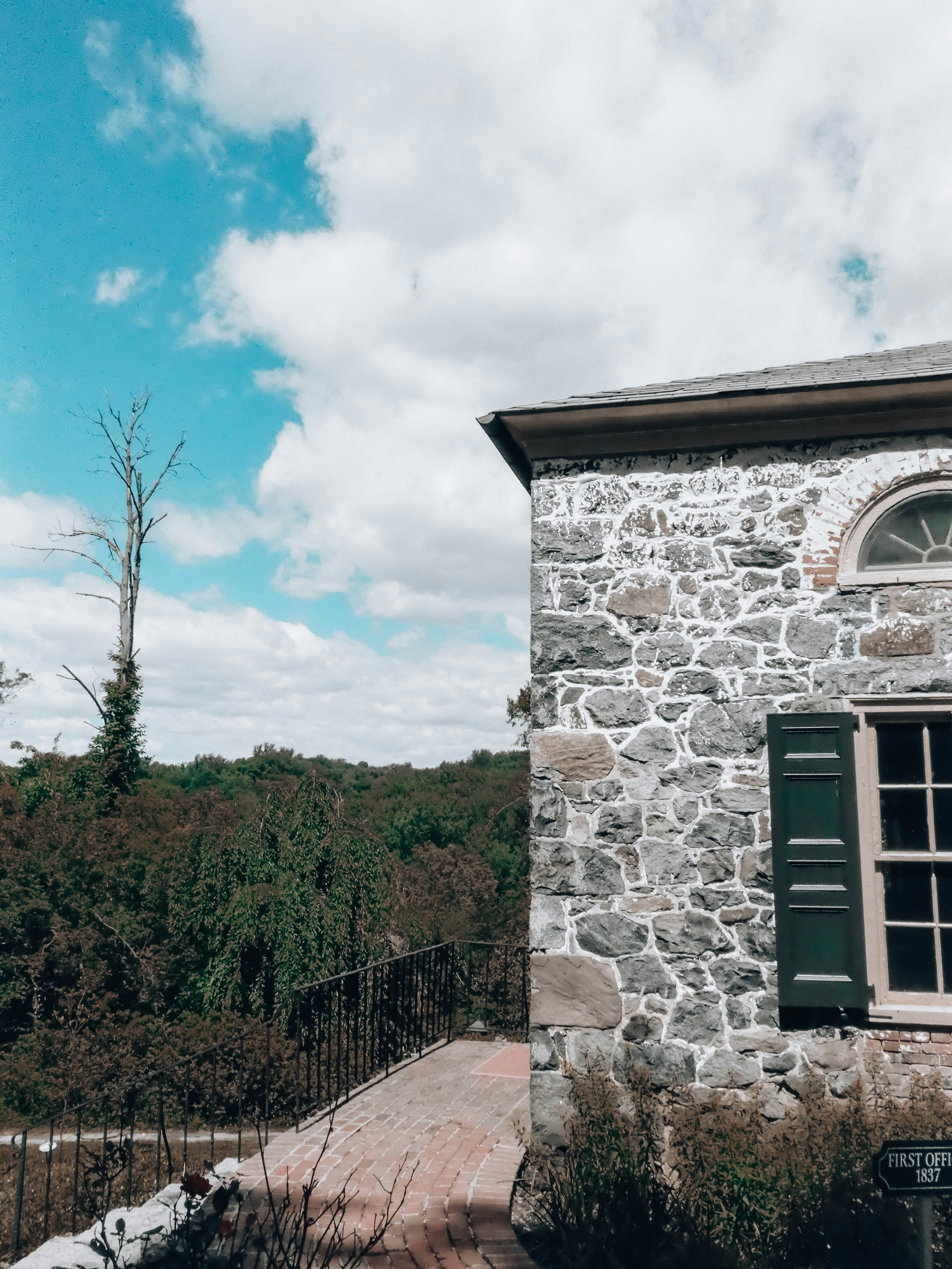 Stone house with arched window and green shutters, set beside a brick path and garden under a bright blue sky.