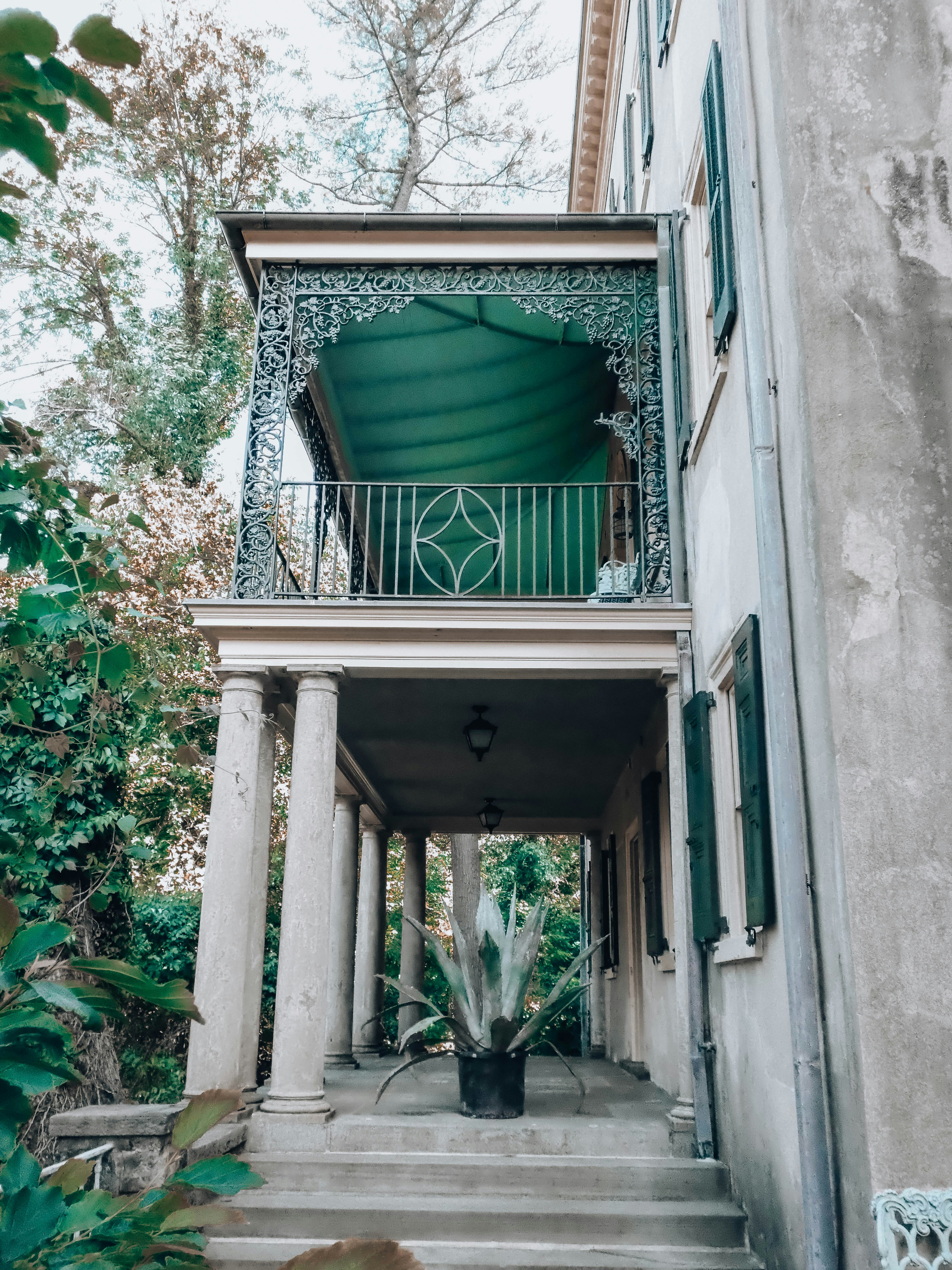 Old-world veranda with intricate ironwork above a shaded stone staircase and potted plant, framed by greenery along a weathered residence.