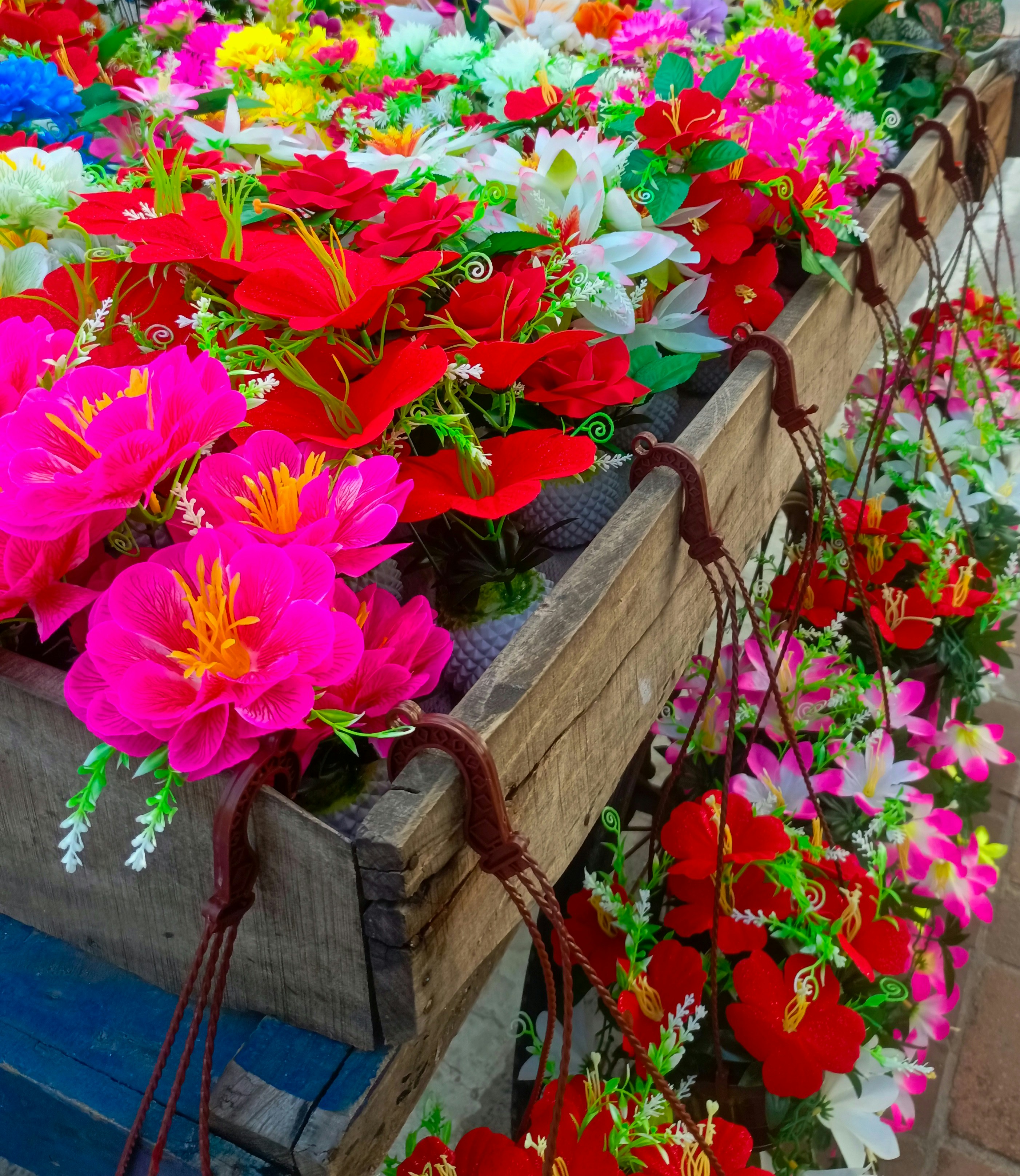 Bright paper flowers spill from a rustic wooden basket, creating a vivid cascade of reds, pinks, and yellows with green foliage and trailing stems.