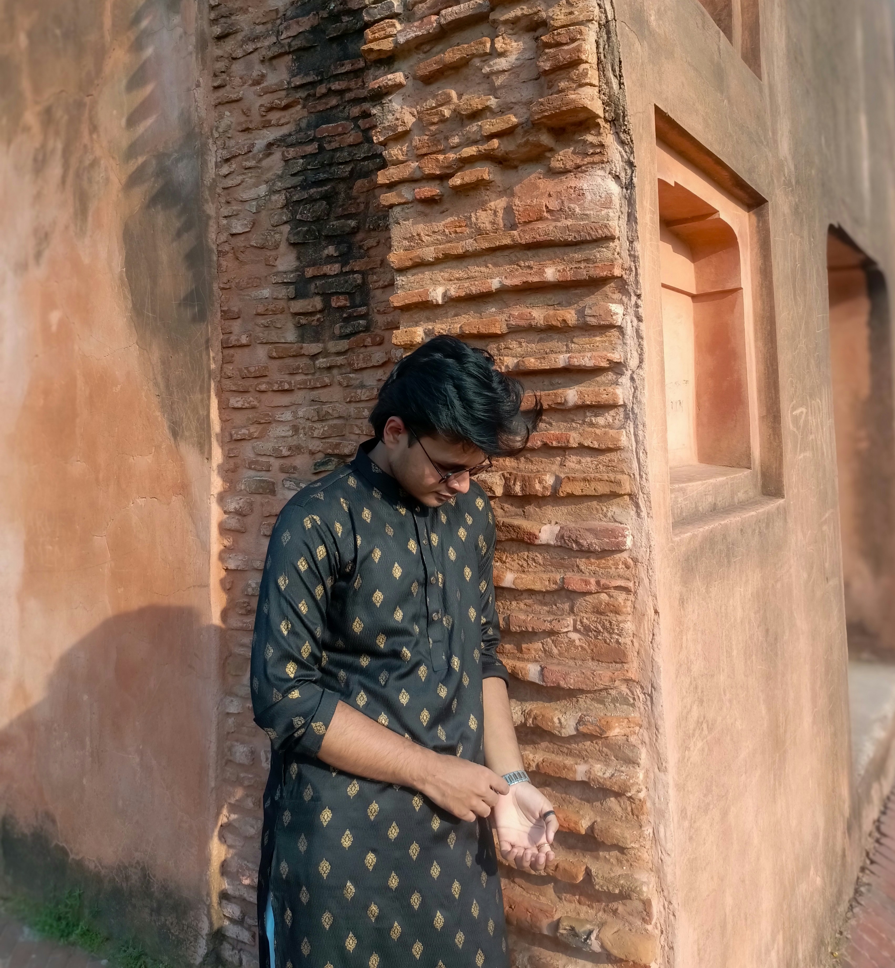 A man in a patterned kurta stands beside a brick wall and an arched window, hands cupped as if adjusting a small object; warm, sunlit tones emphasize textures of the wall and clothing.