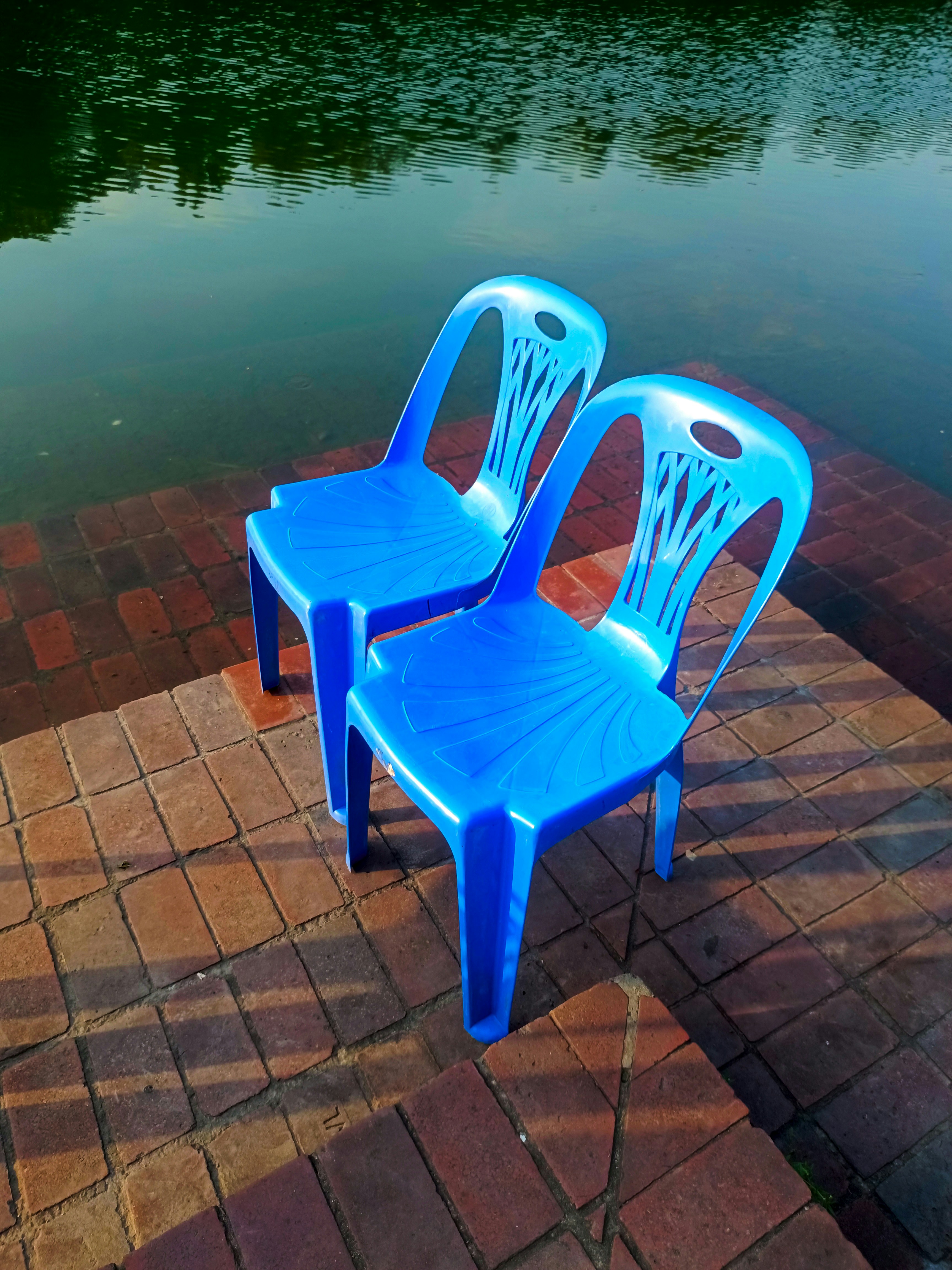 Two vibrant blue plastic chairs sit on a brick-paved edge beside calm water, inviting a quiet moment by the riverbank.