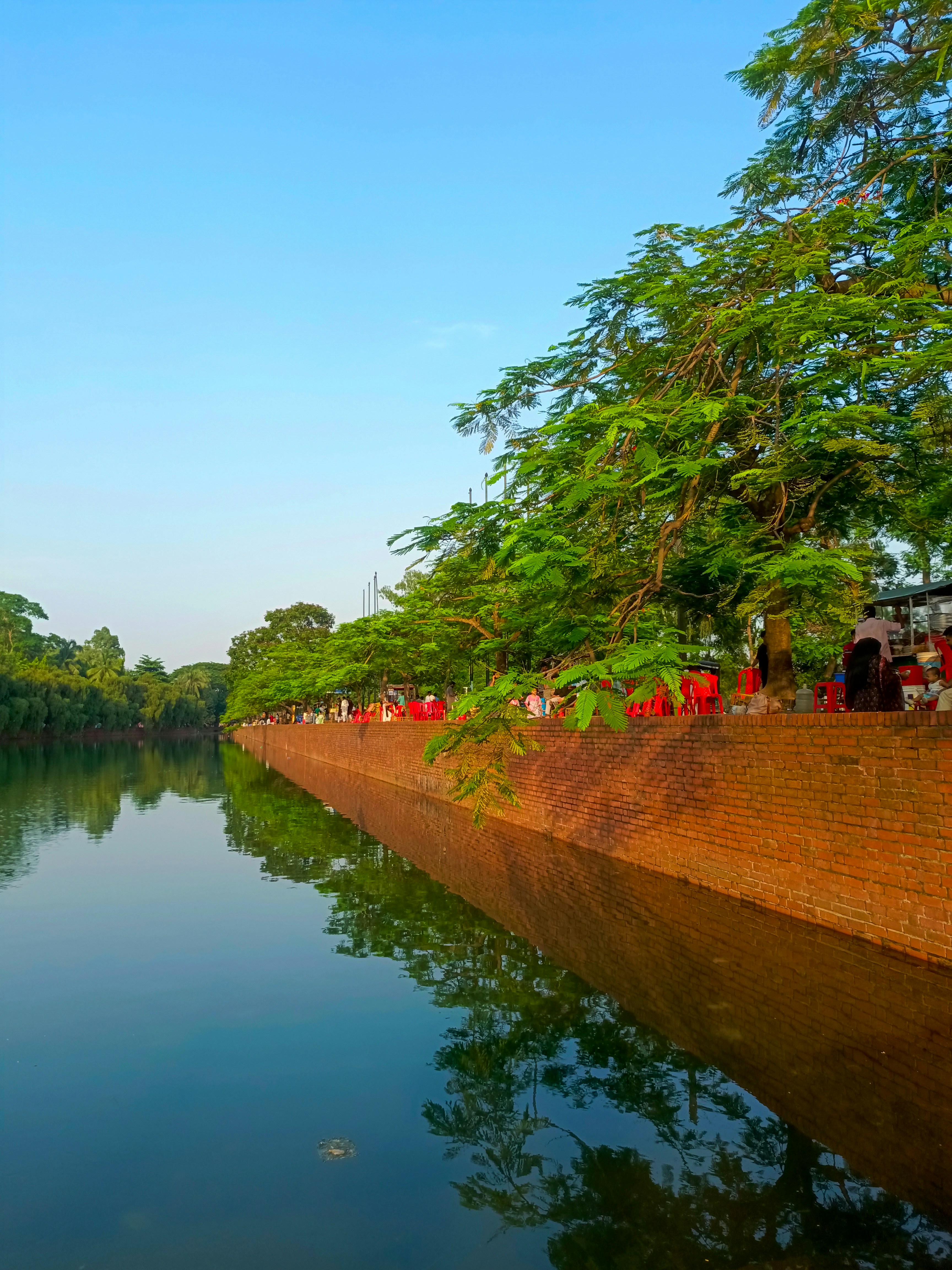 Calm canal reflects blue sky, brick wall, and green trees.