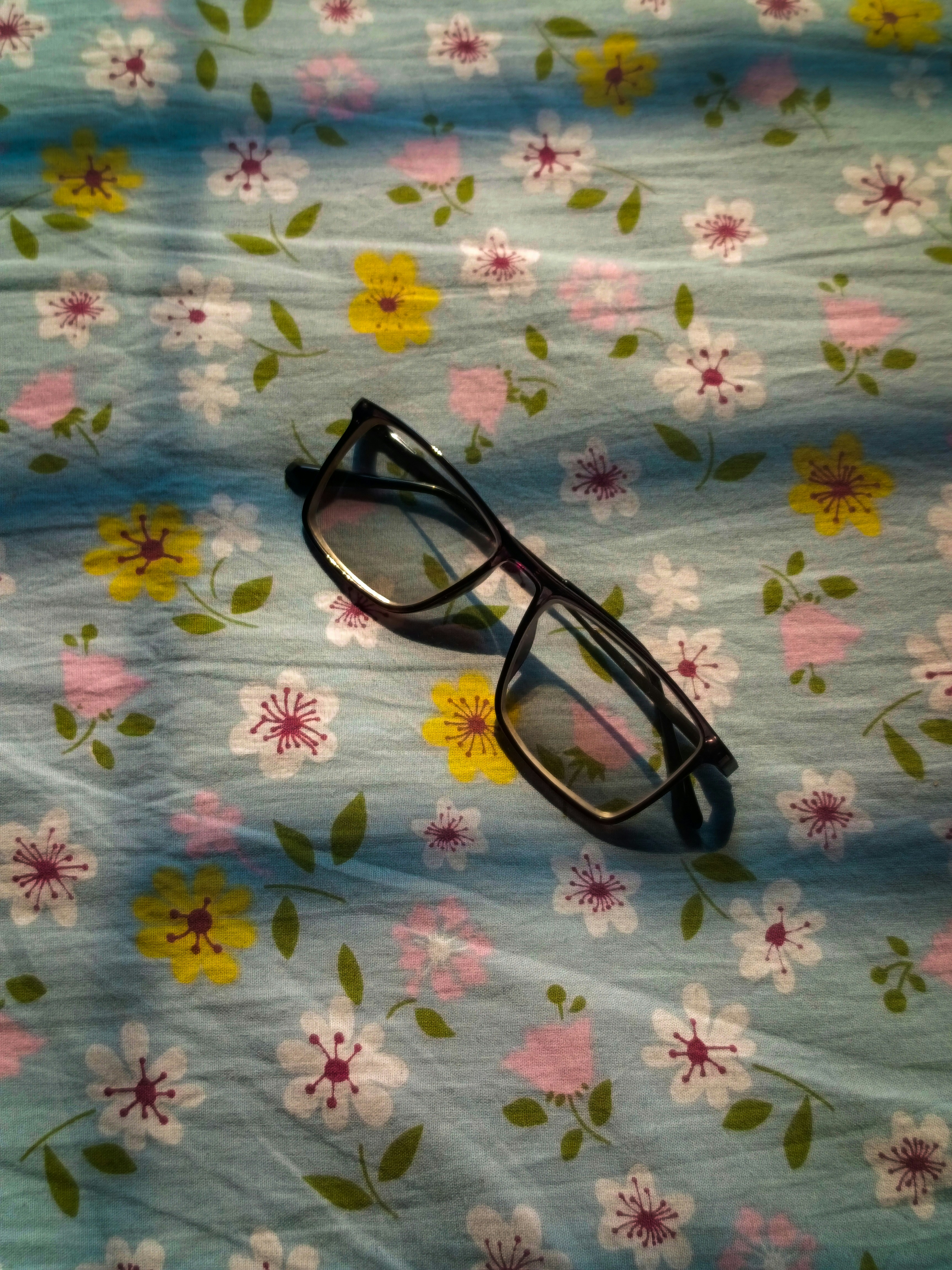 Pair of black eyeglasses rests on a pastel floral bedsheet, softly lit and centered to emphasize everyday detail.