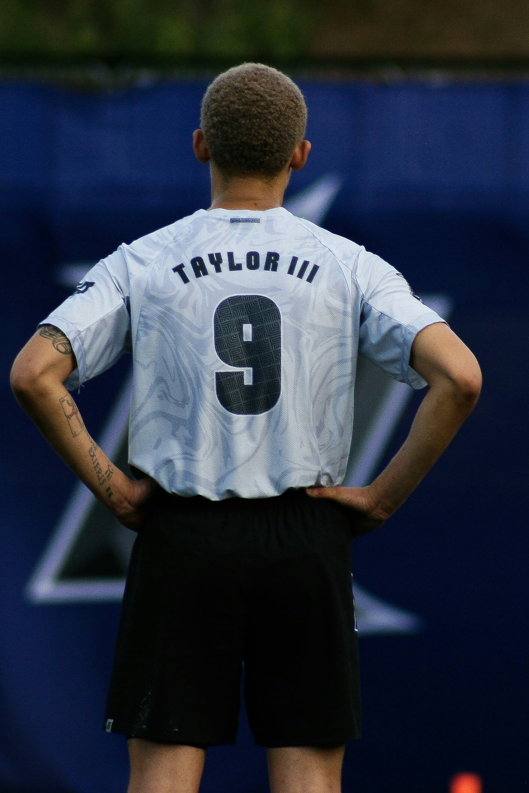 Soccer player seen from behind wearing a light jersey with the name and number 9. The focus is on the athlete standing with hands on hips against a dark backdrop.