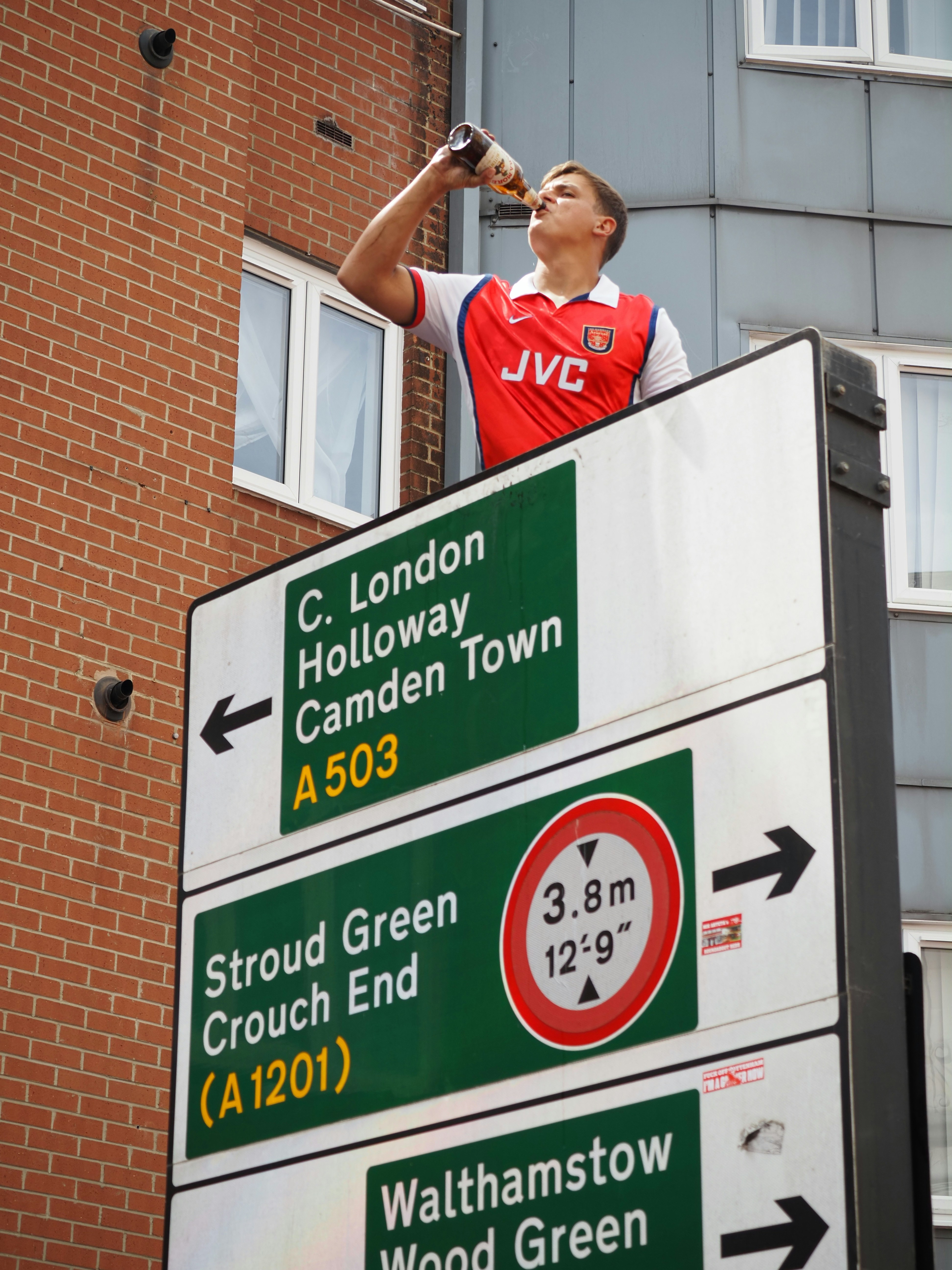 Arms Raised atop a City Sign: A Man in a Red Arsenal Jersey Quenching Midday Heat