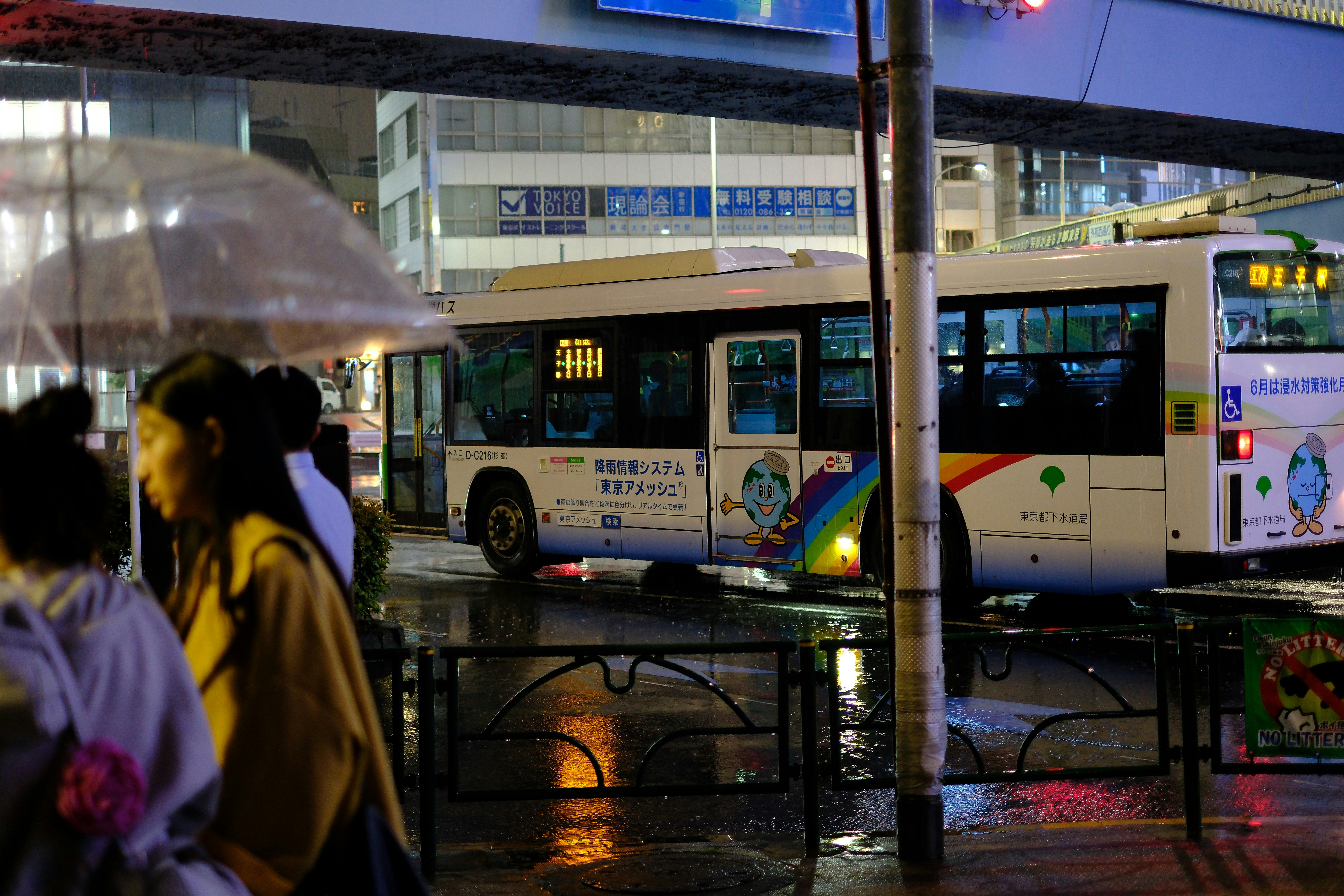 A city bus passes through Tokyo at night in the rain, with glowing streetlights and neon reflections creating a moody urban scene.