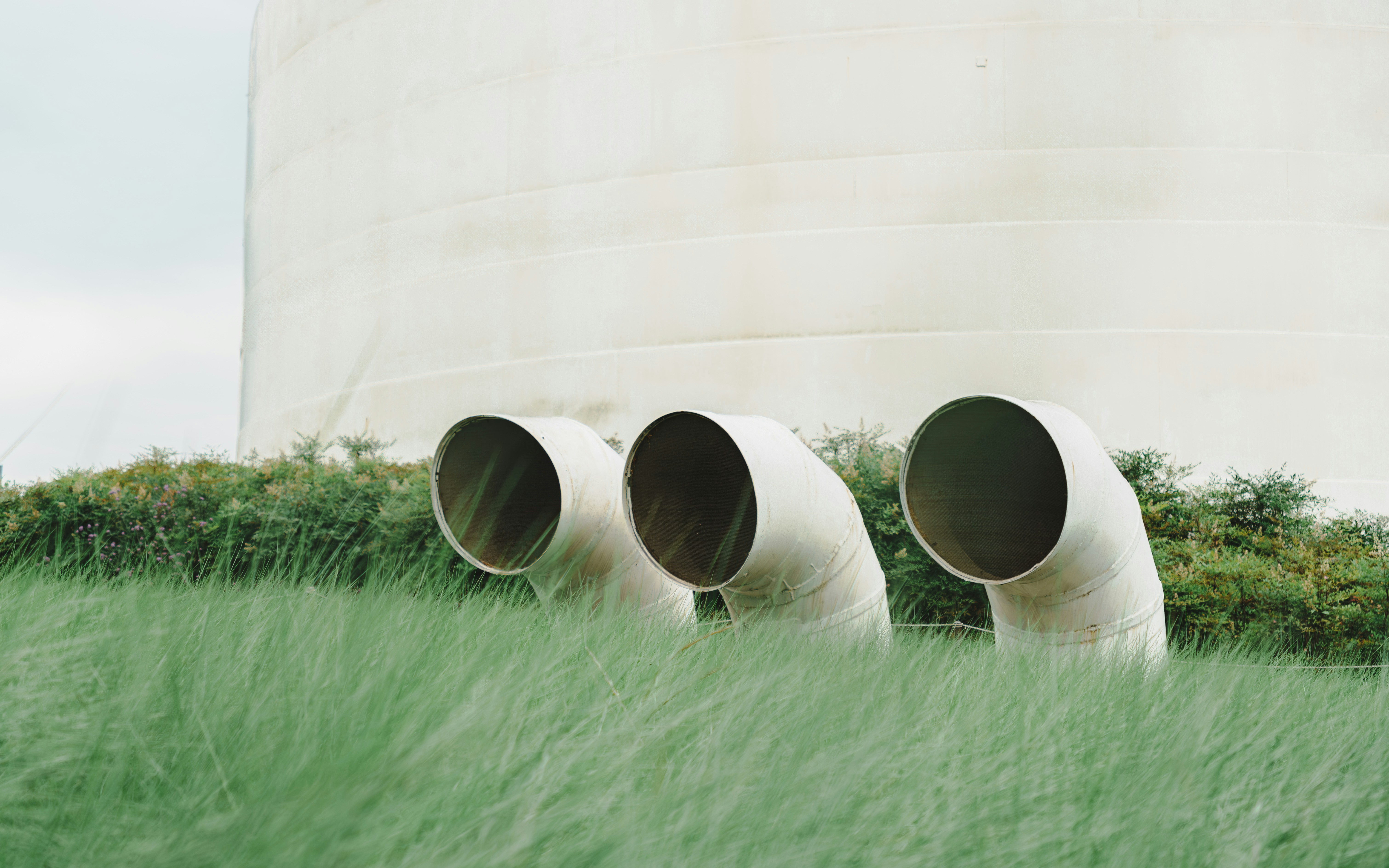 Three industrial pipes piercing a grassy field beneath a pale silo