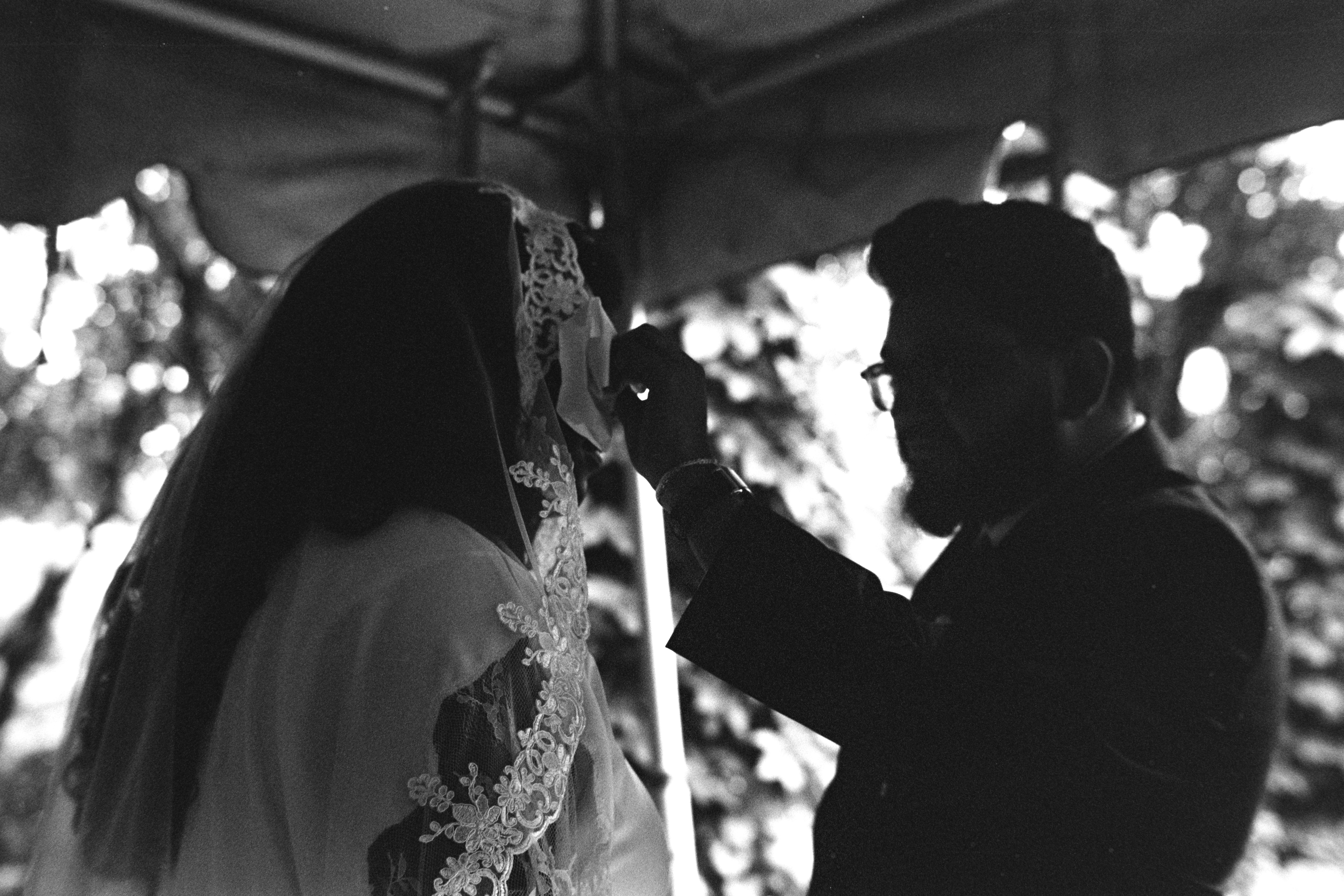 Silhouetted man adjusts bride's veil outdoors.