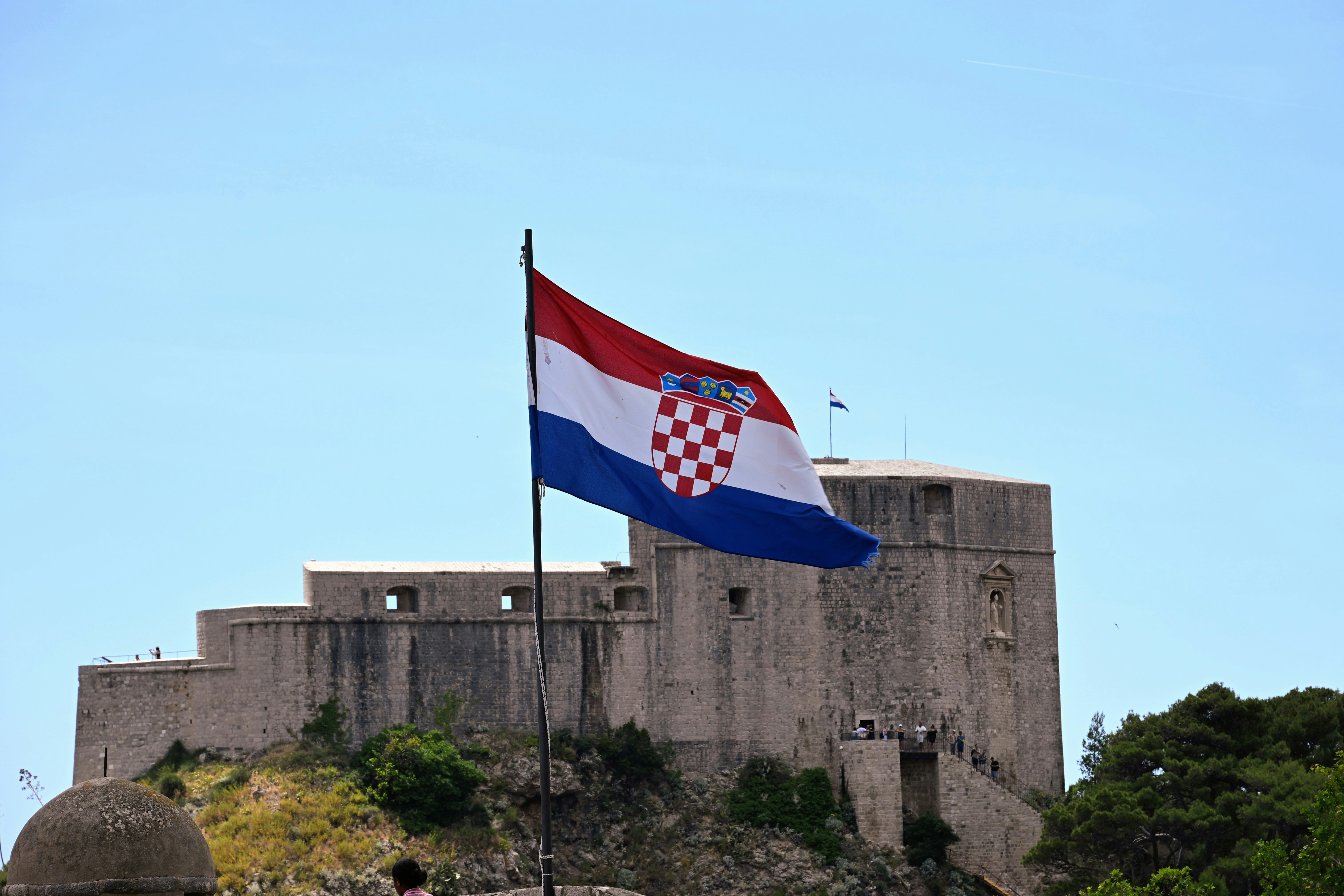The ancient walls of Dubrovnik trace the edge of a rugged limestone cliff, overlooking the deep blue waters of the Adriatic Sea on Croatia's southern coast.