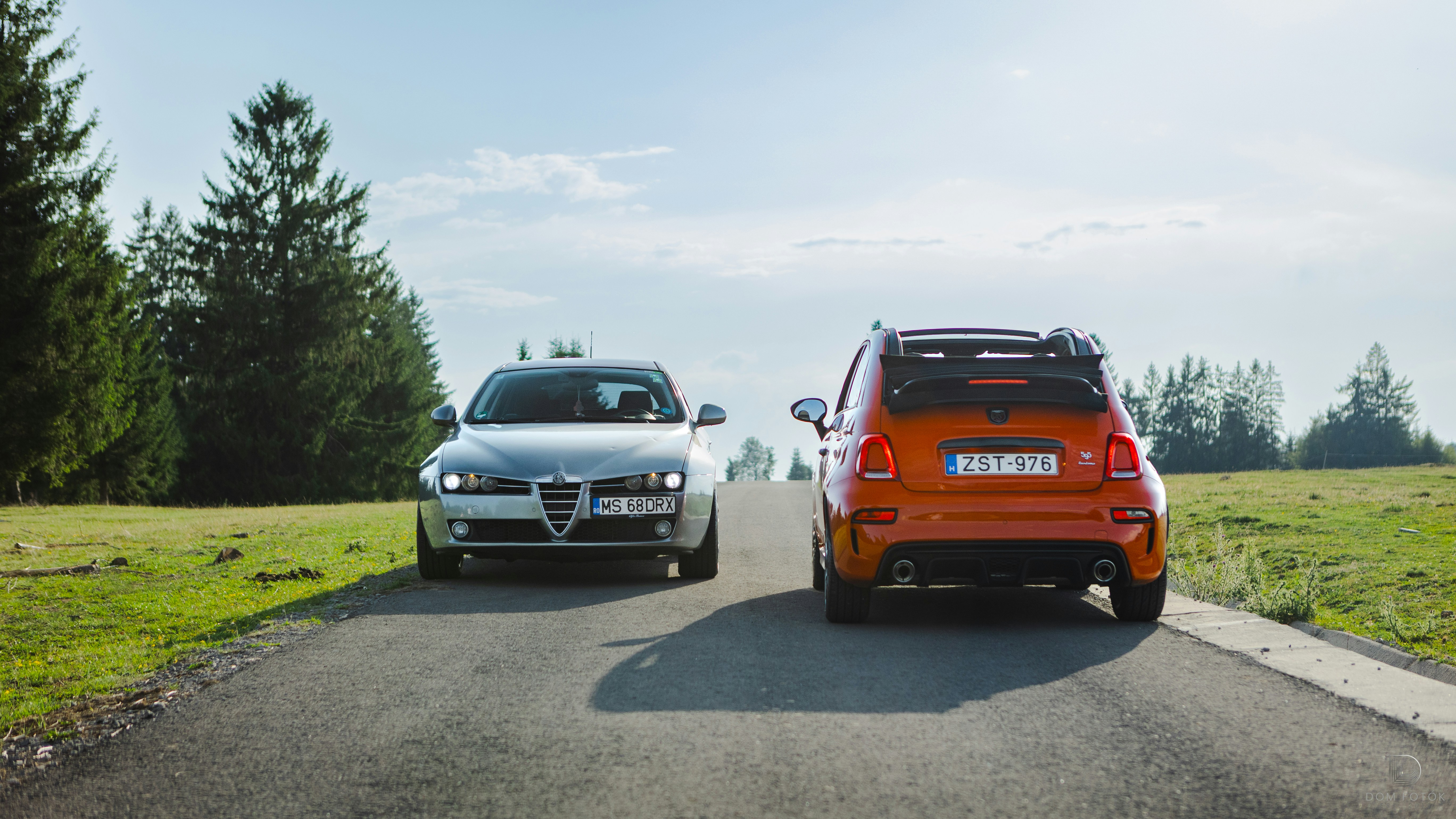 Head-to-head on the rural road: silver sedan and orange hatch confront the horizon