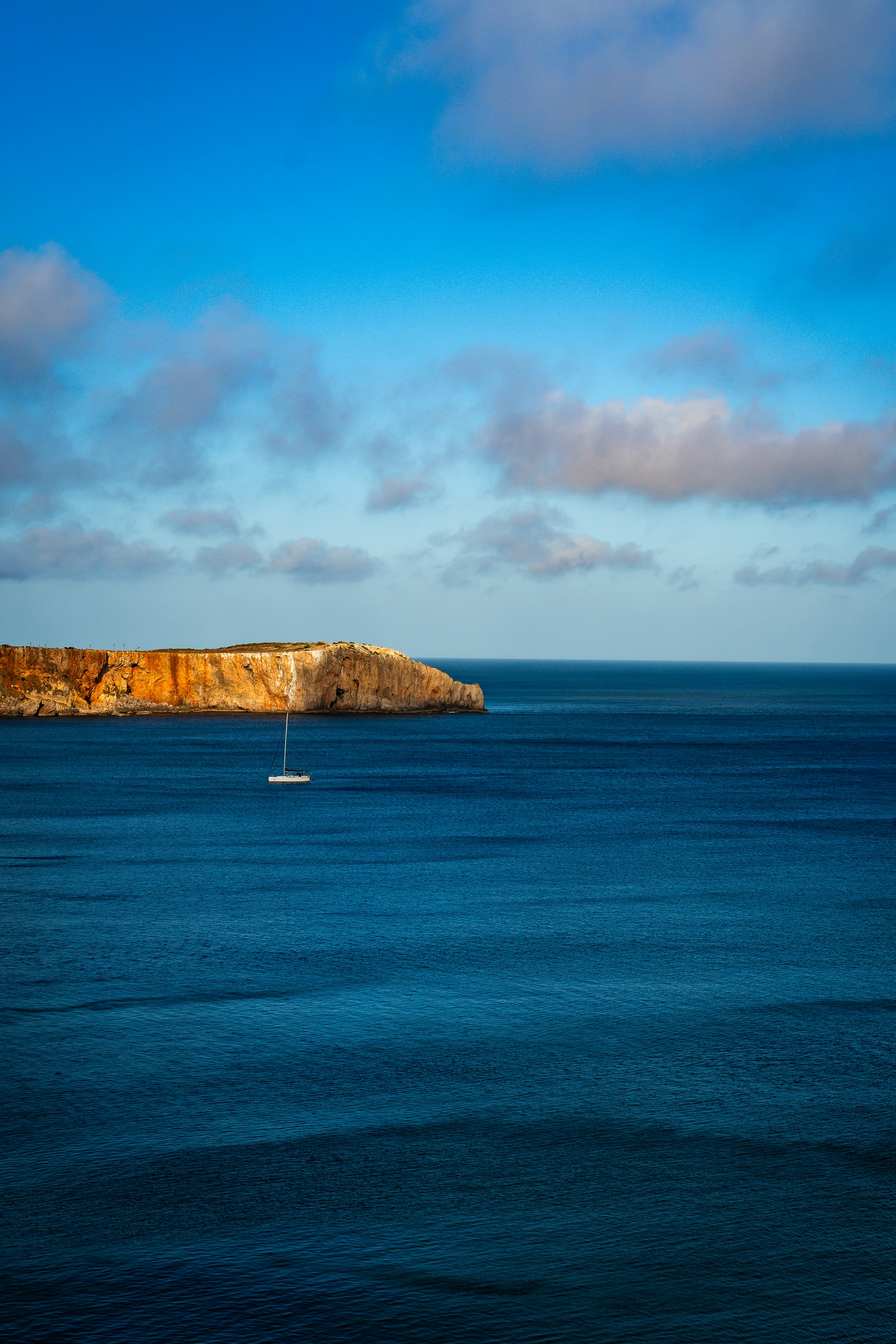 White sailboat near sunlit cliff on a calm blue sea
