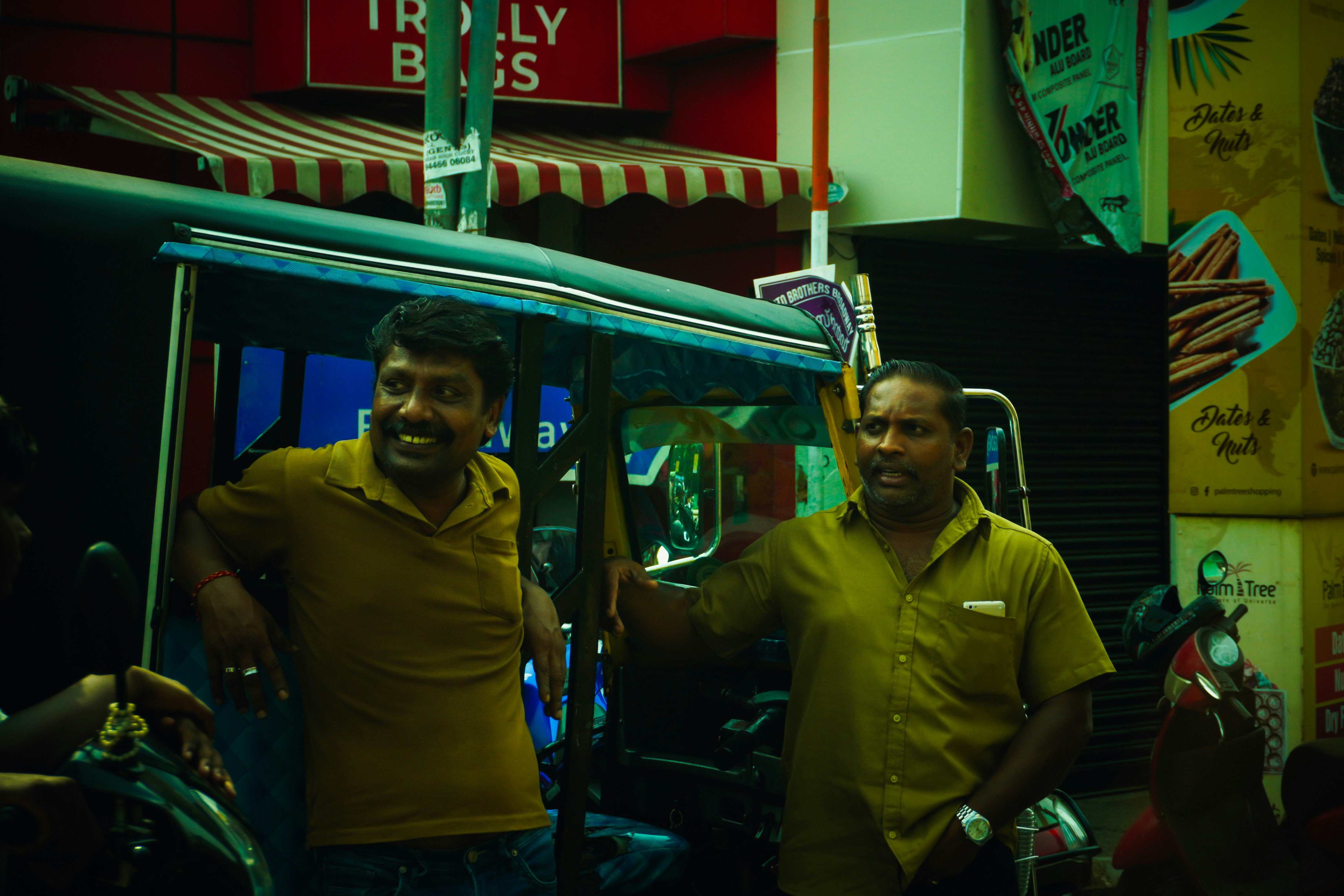 Two street vendors in olive shirts stand by a parked auto-rickshaw amid a nighttime market glow