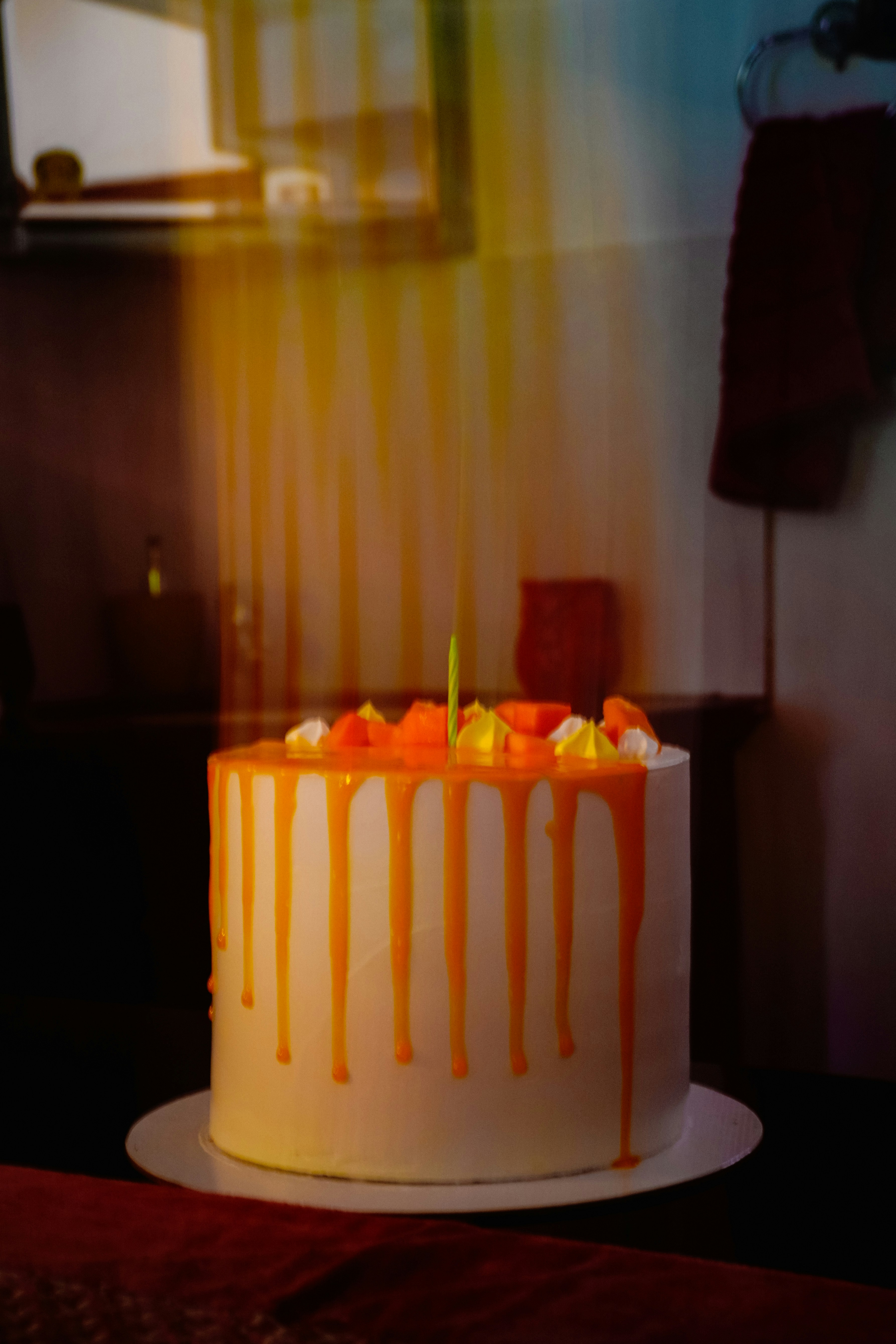 A white cake with orange icing and a lit candle.