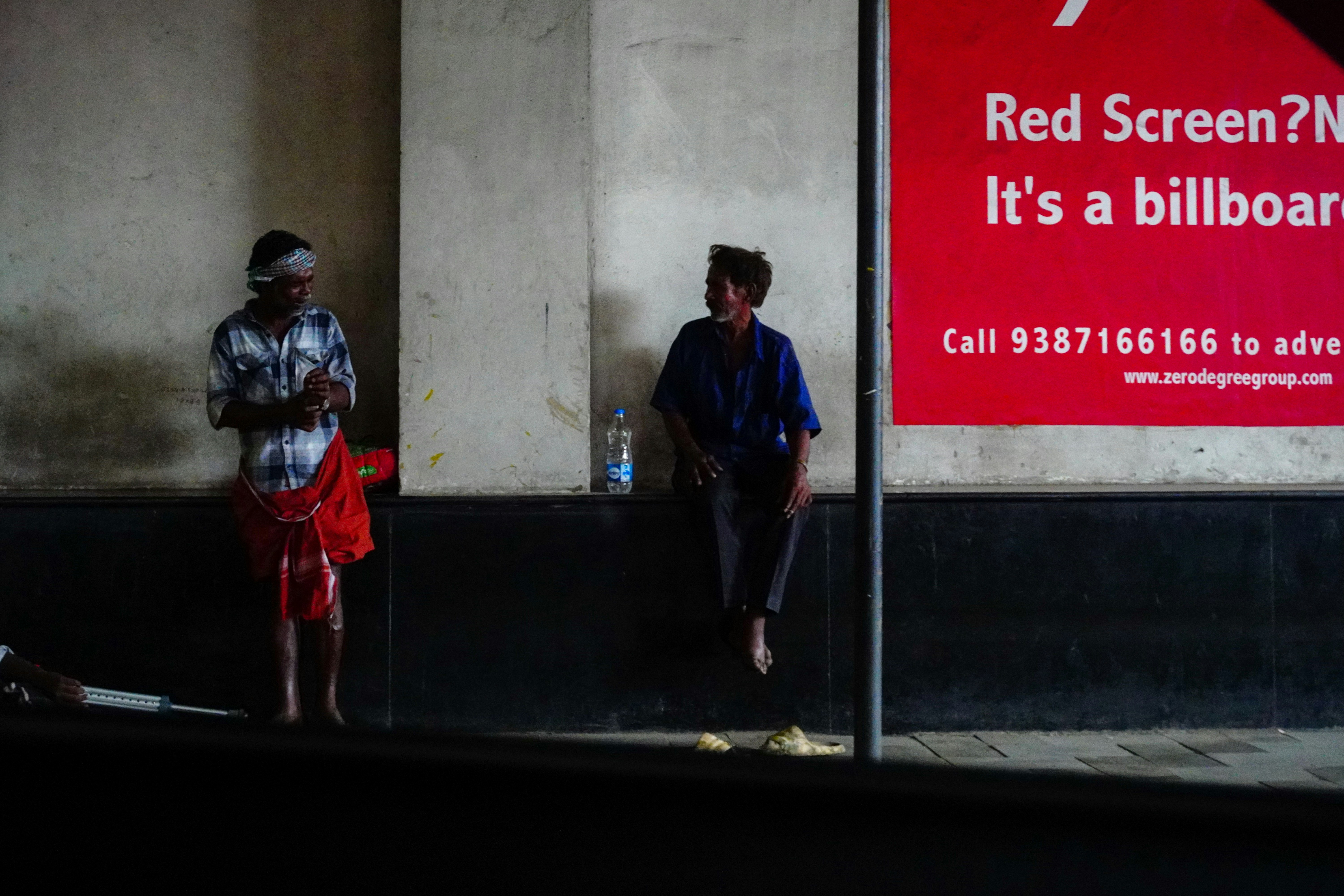 Two people stand near a red billboard in dim light.