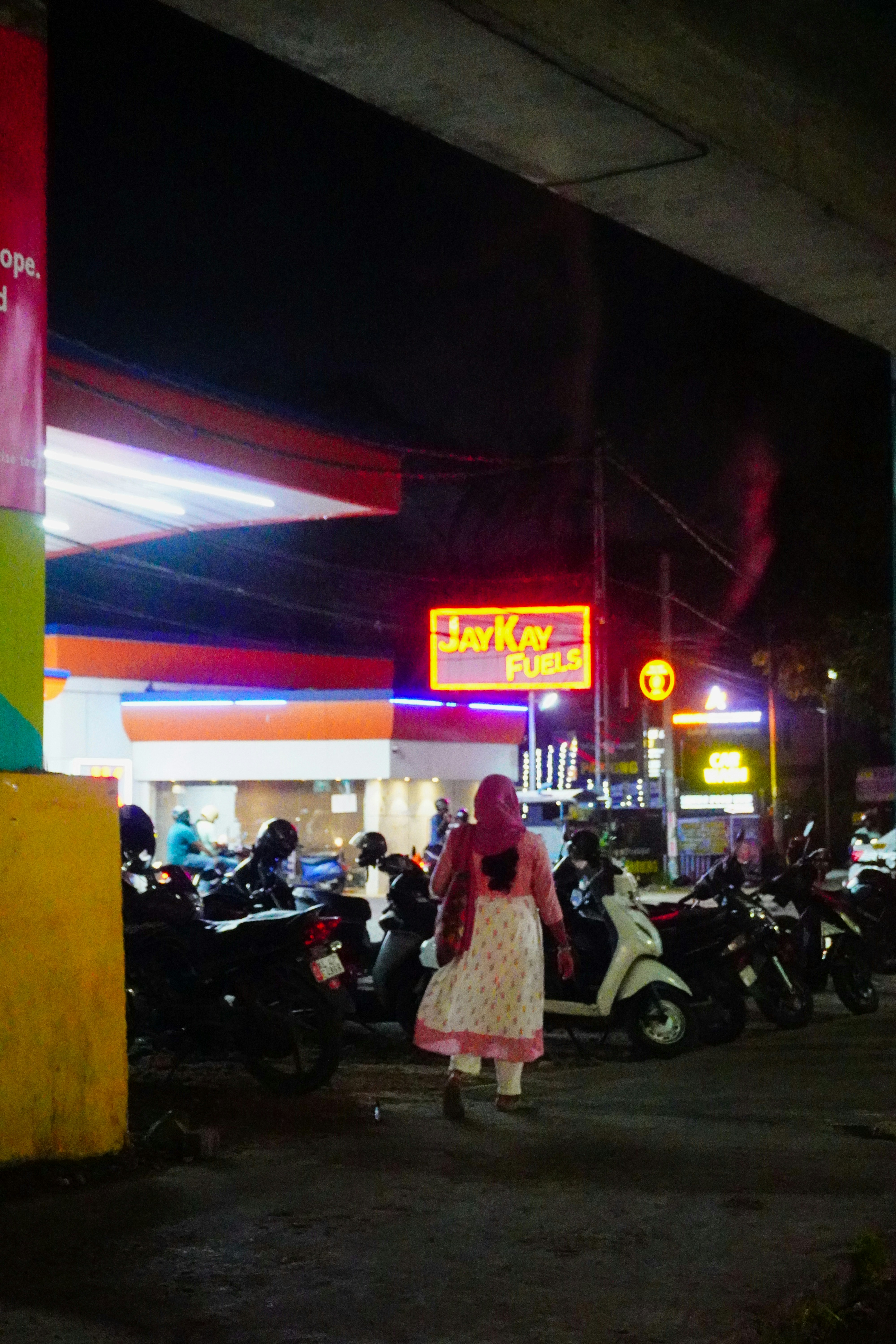 Woman walks past parked motorcycles near a lit fuel station.