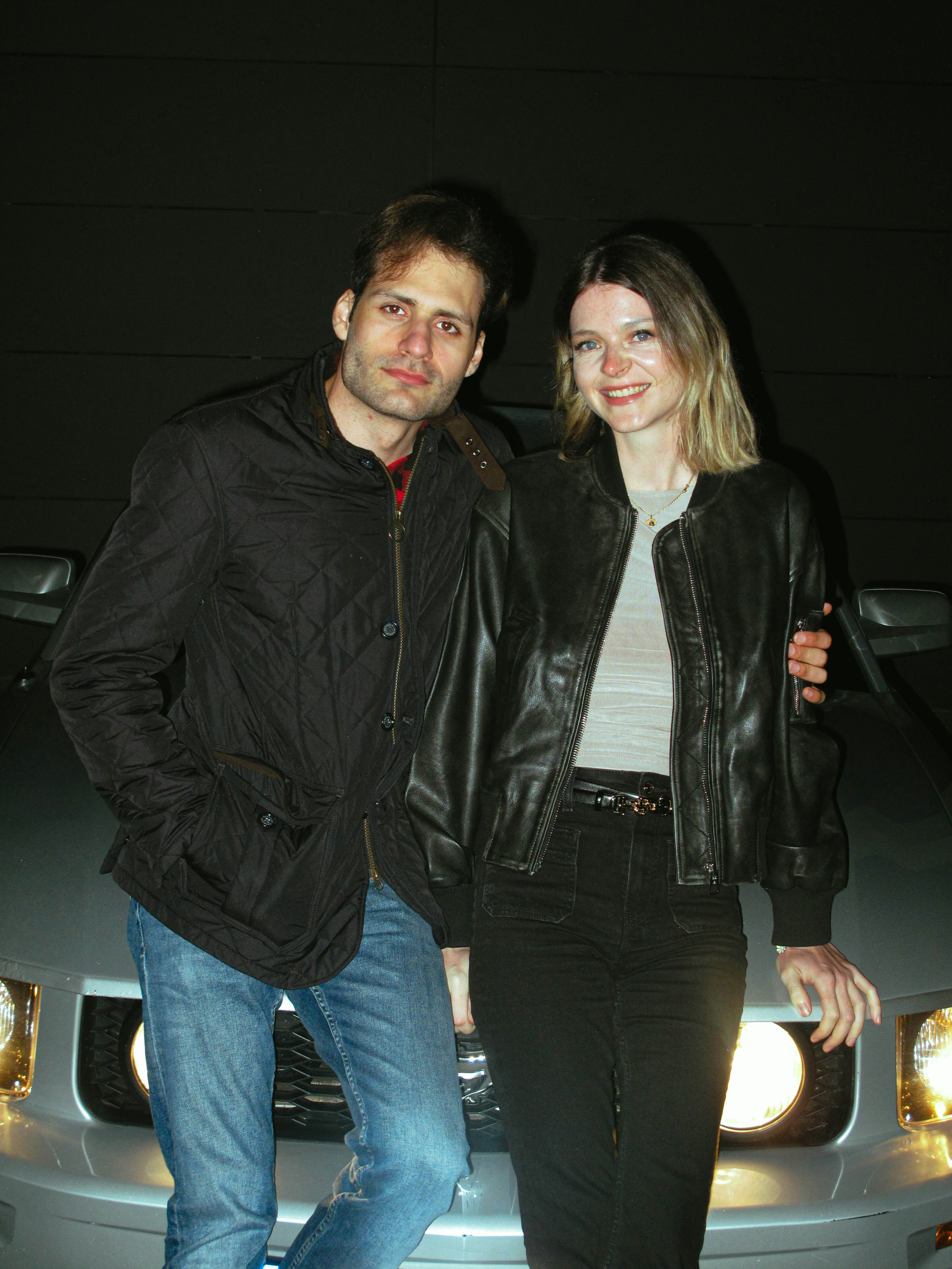 Two people pose closely in front of a car at night, wearing black leather jackets; headlights provide backlight and the scene emphasizes casual companionship.
