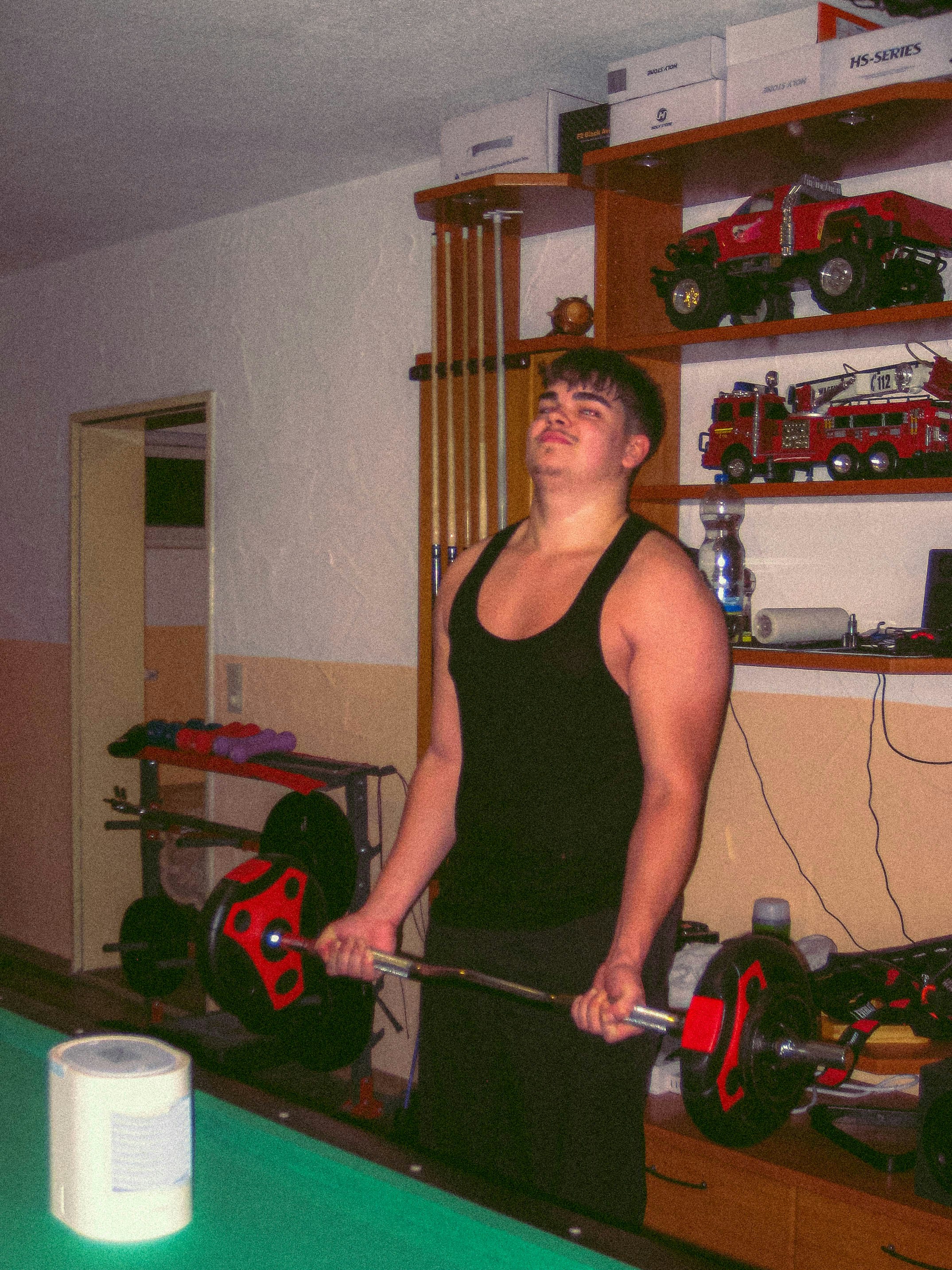 Athlete lifts a barbell in a cluttered basement gym with shelves of models and boxes in the background.