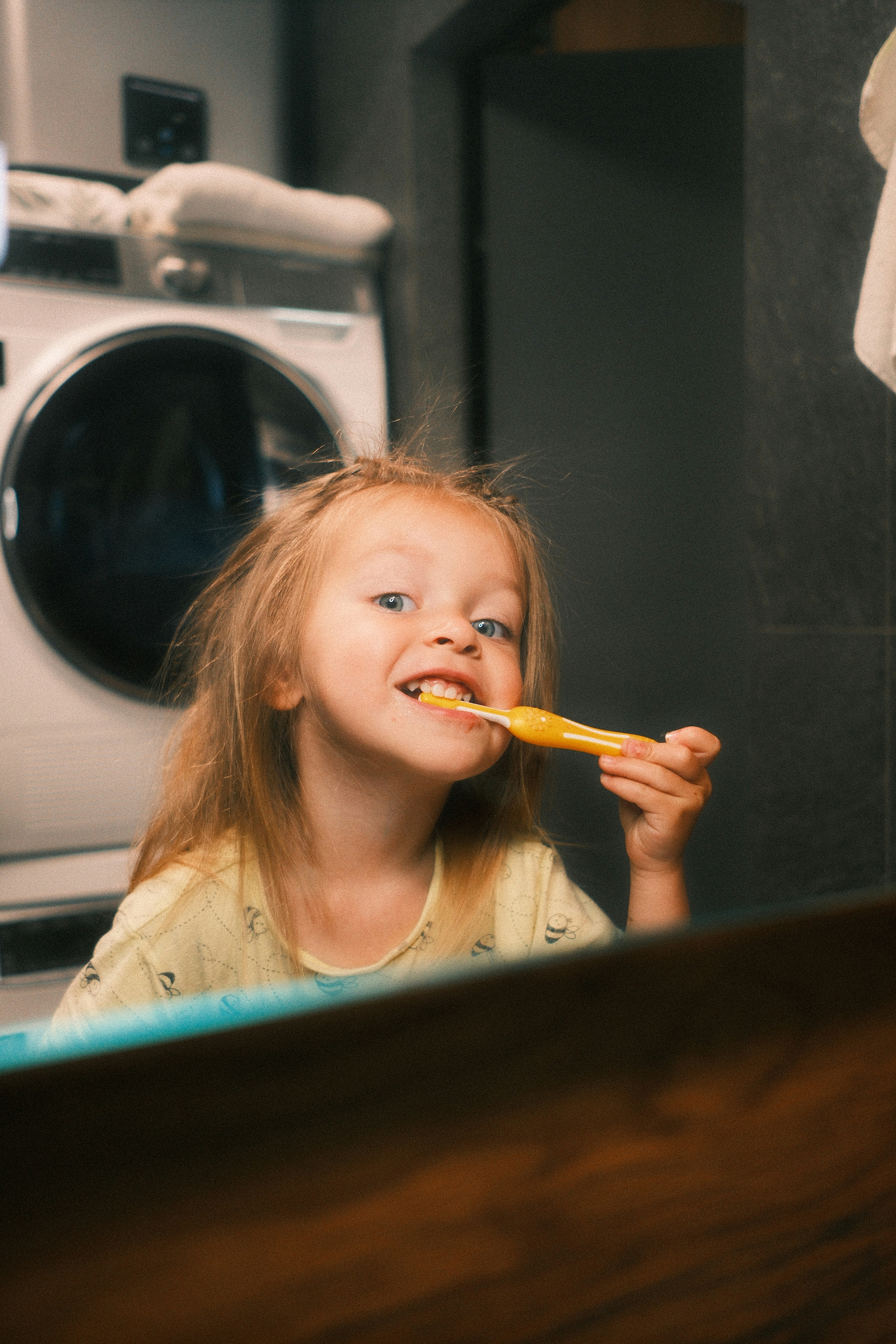 A young girl brushes her teeth in the bathroom mirror.
