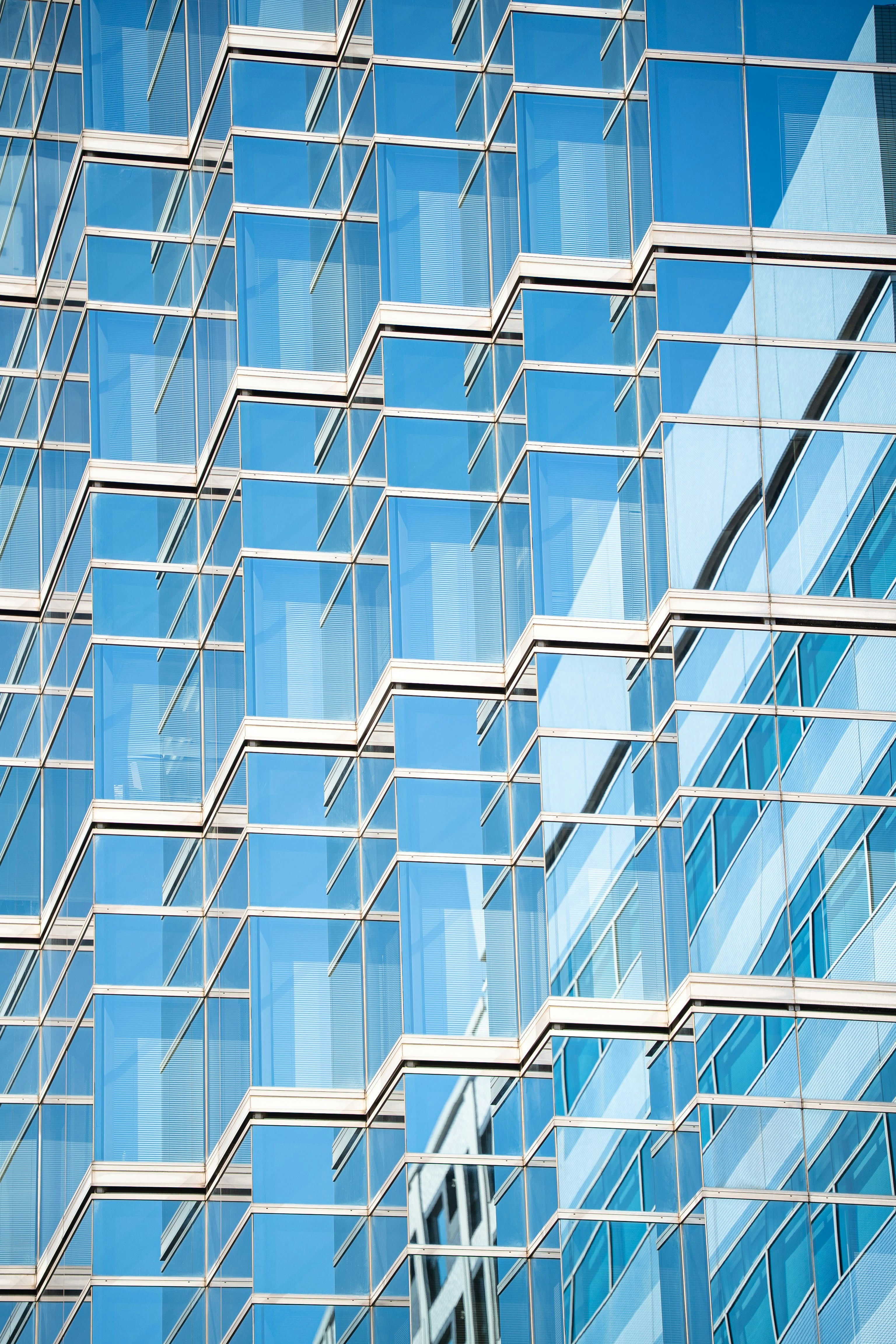 Abstract close-up of a modern glass building facade reflecting the blue sky. Repetitive geometric patterns of steel frames and glass panels create a rhythmic, grid-like composition.