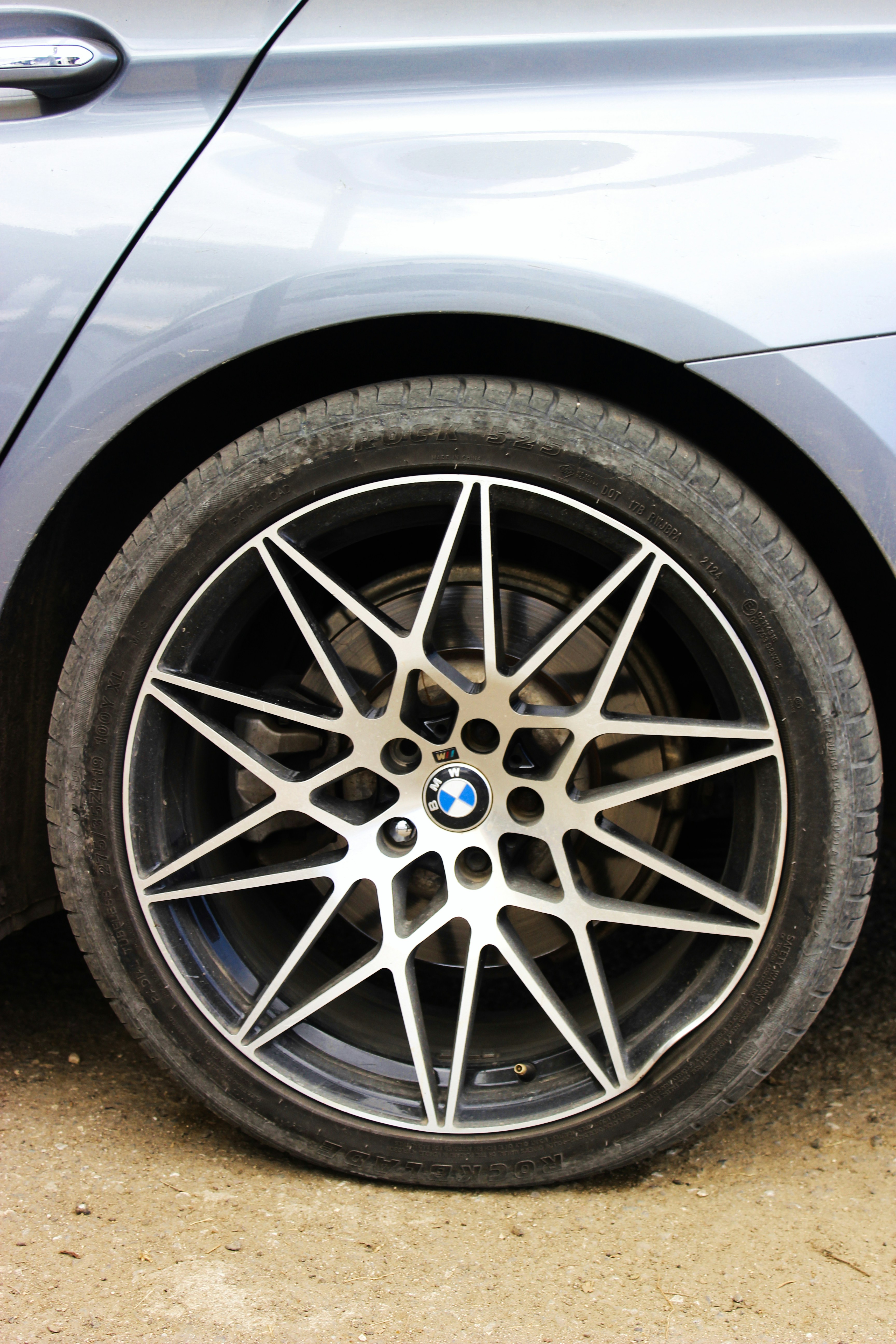 A close-up shot of a modern BMW multi-spoke alloy rim, showcasing the intricate silver and black star pattern design, brake disc detailing, and low-profile tire mounted on a grey luxury sedan.