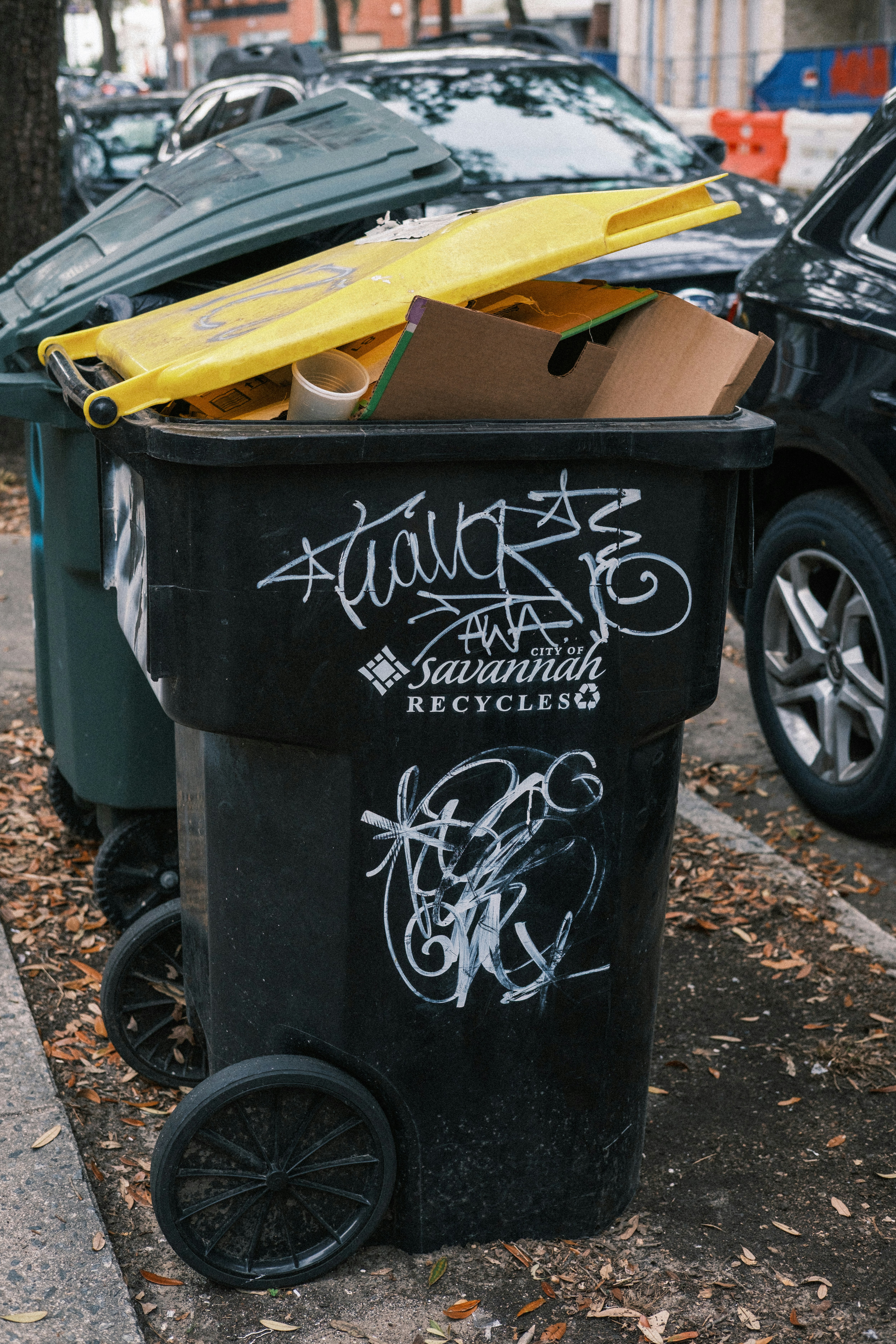 Graffiti-covered black recycling bin overflowing with refuse on street.