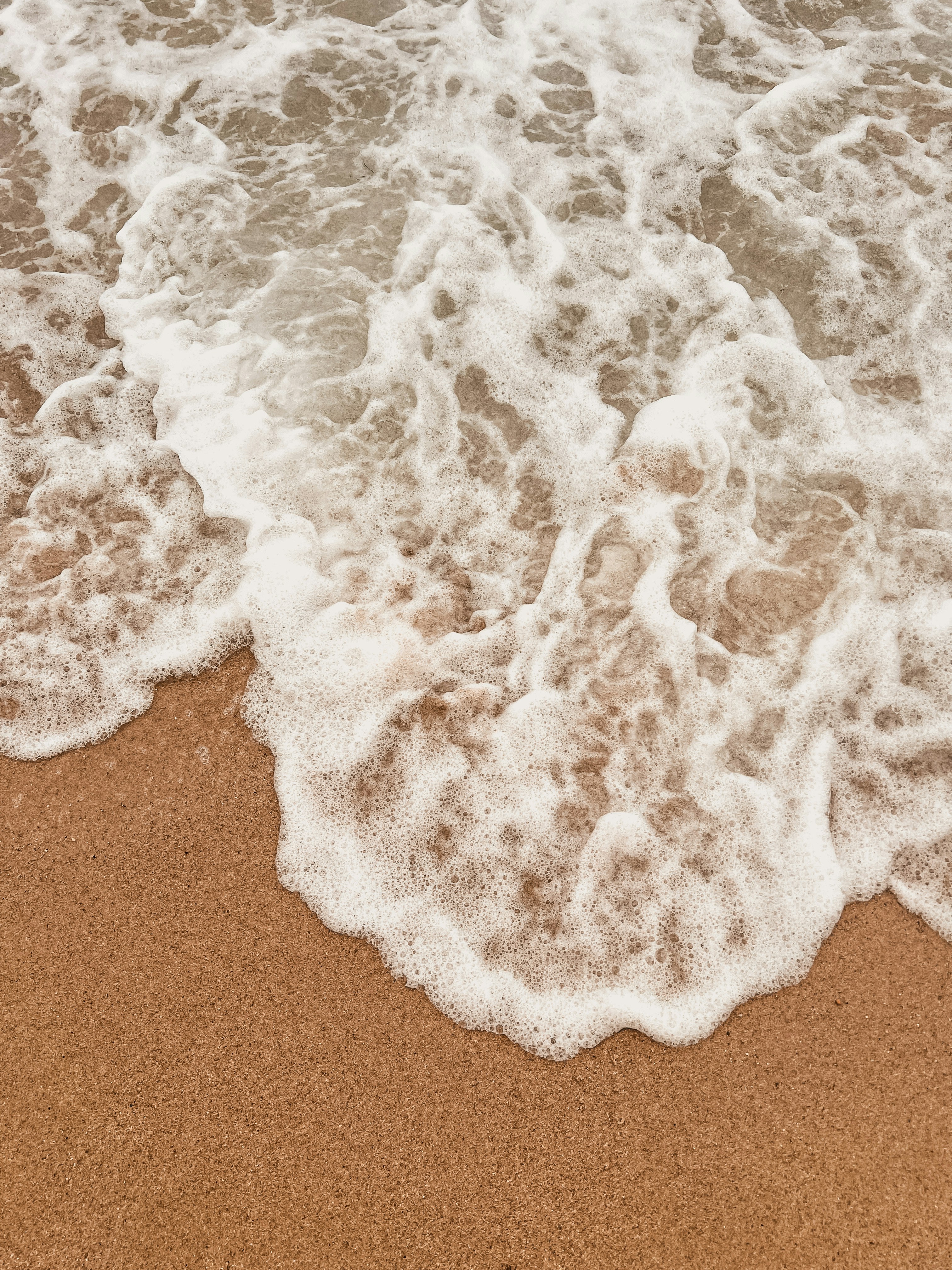Foamy ocean waves wash over a damp sandy beach, capturing the edge where water meets sand in soft, textured swirls.