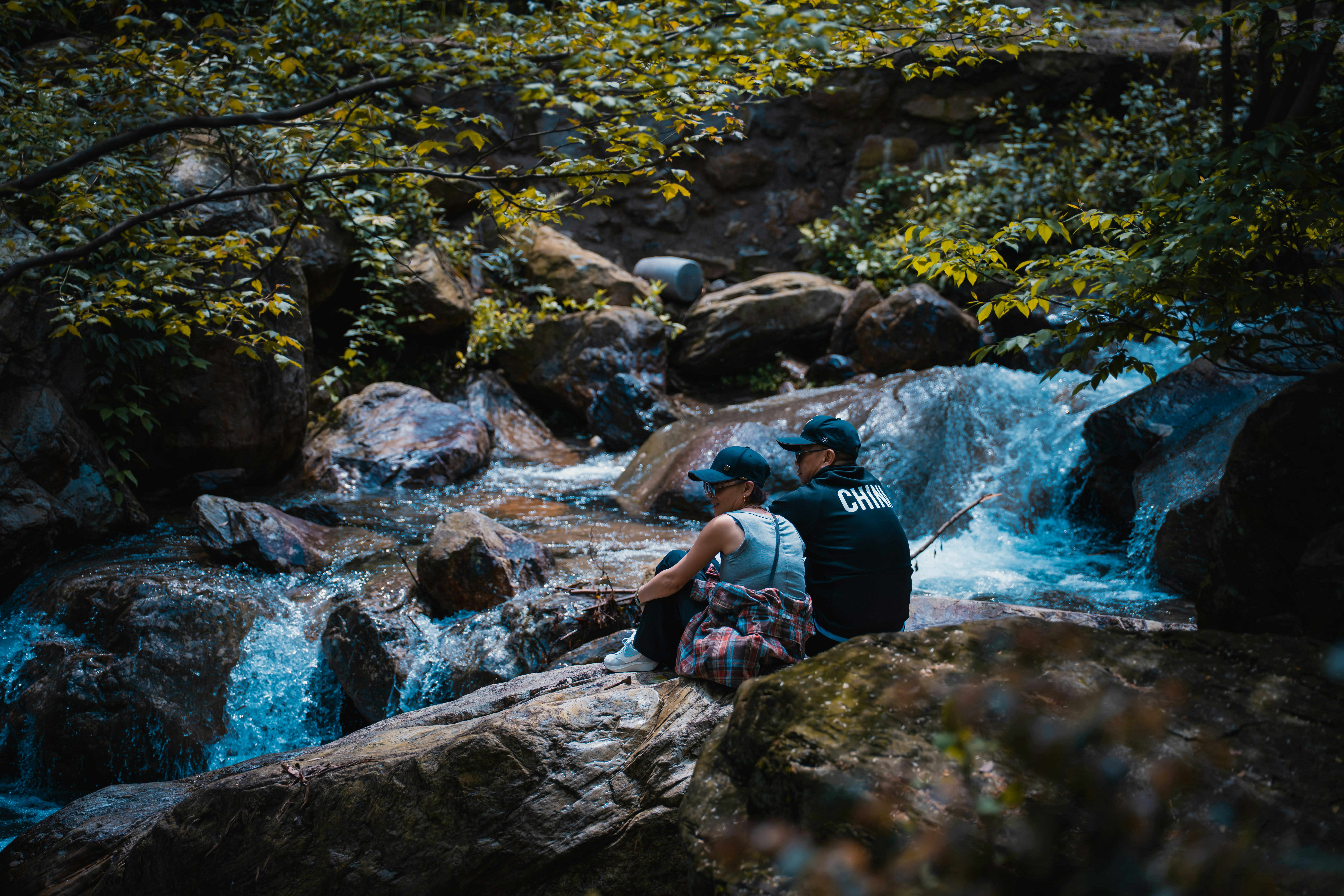 Two hikers pause by a mossy river in a shaded canyon