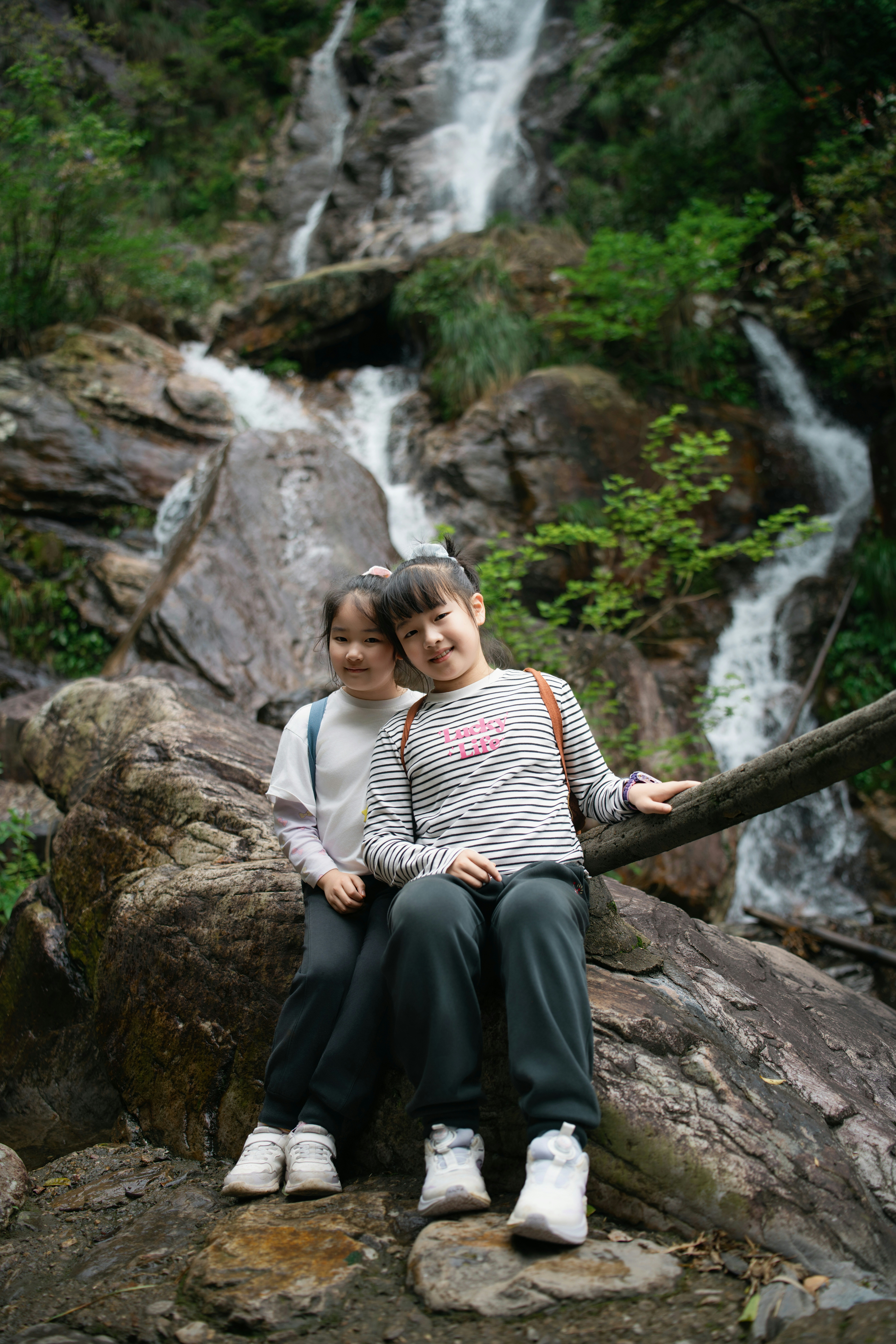Two girls smiling, sitting on rocks near a beautiful waterfall.
