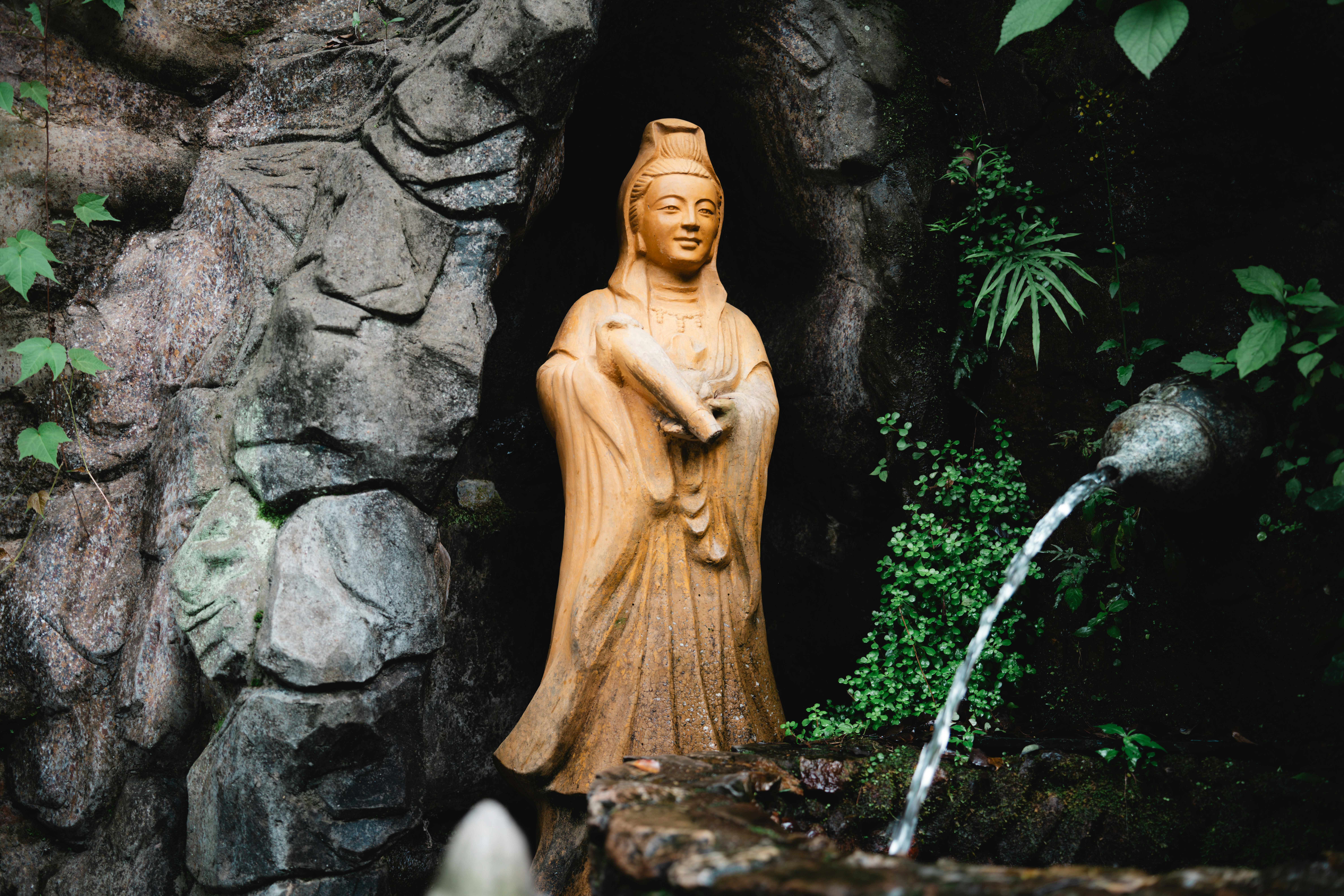 Wooden statue in a grotto with flowing water and greenery.