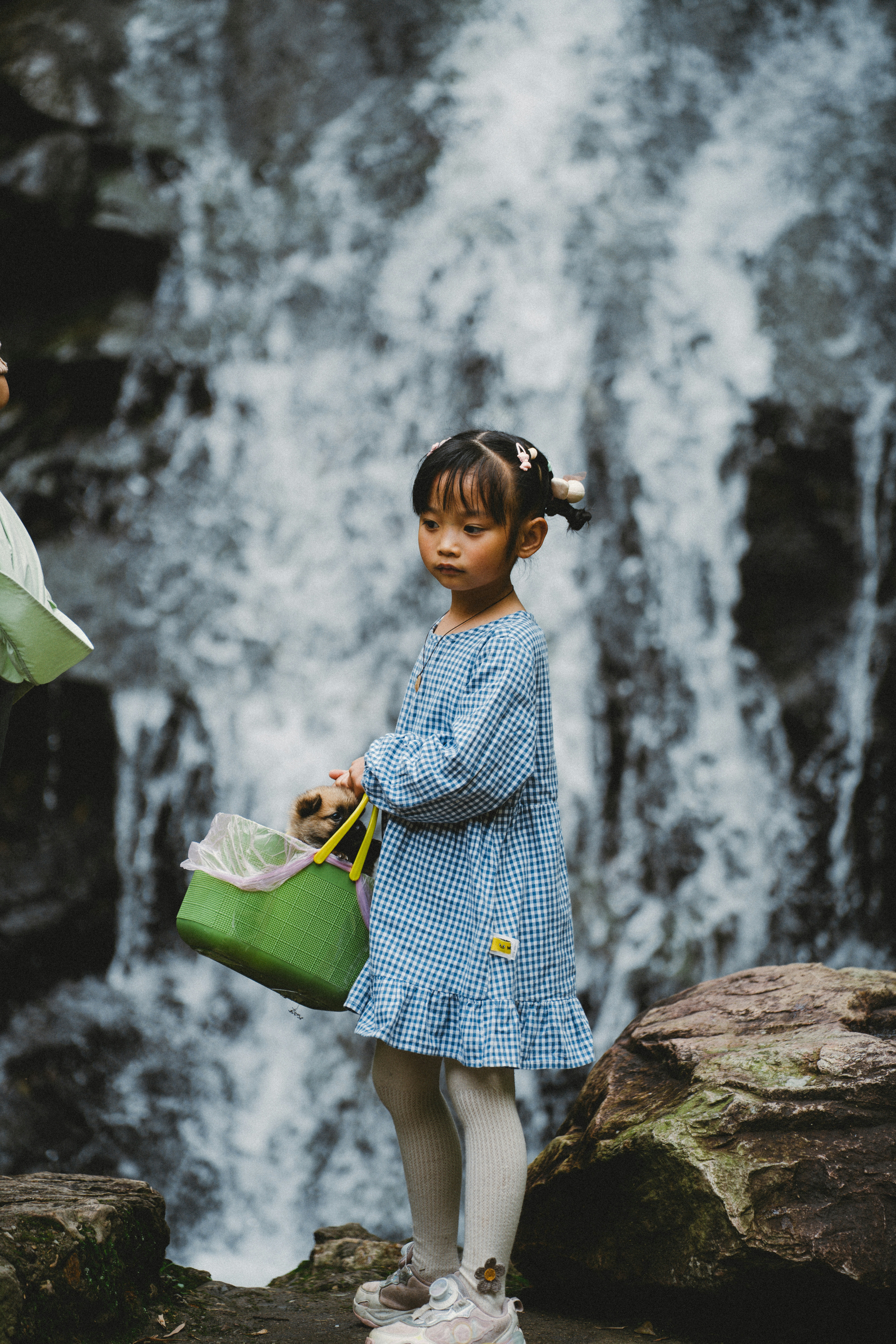 A young girl holds a green basket near a waterfall.