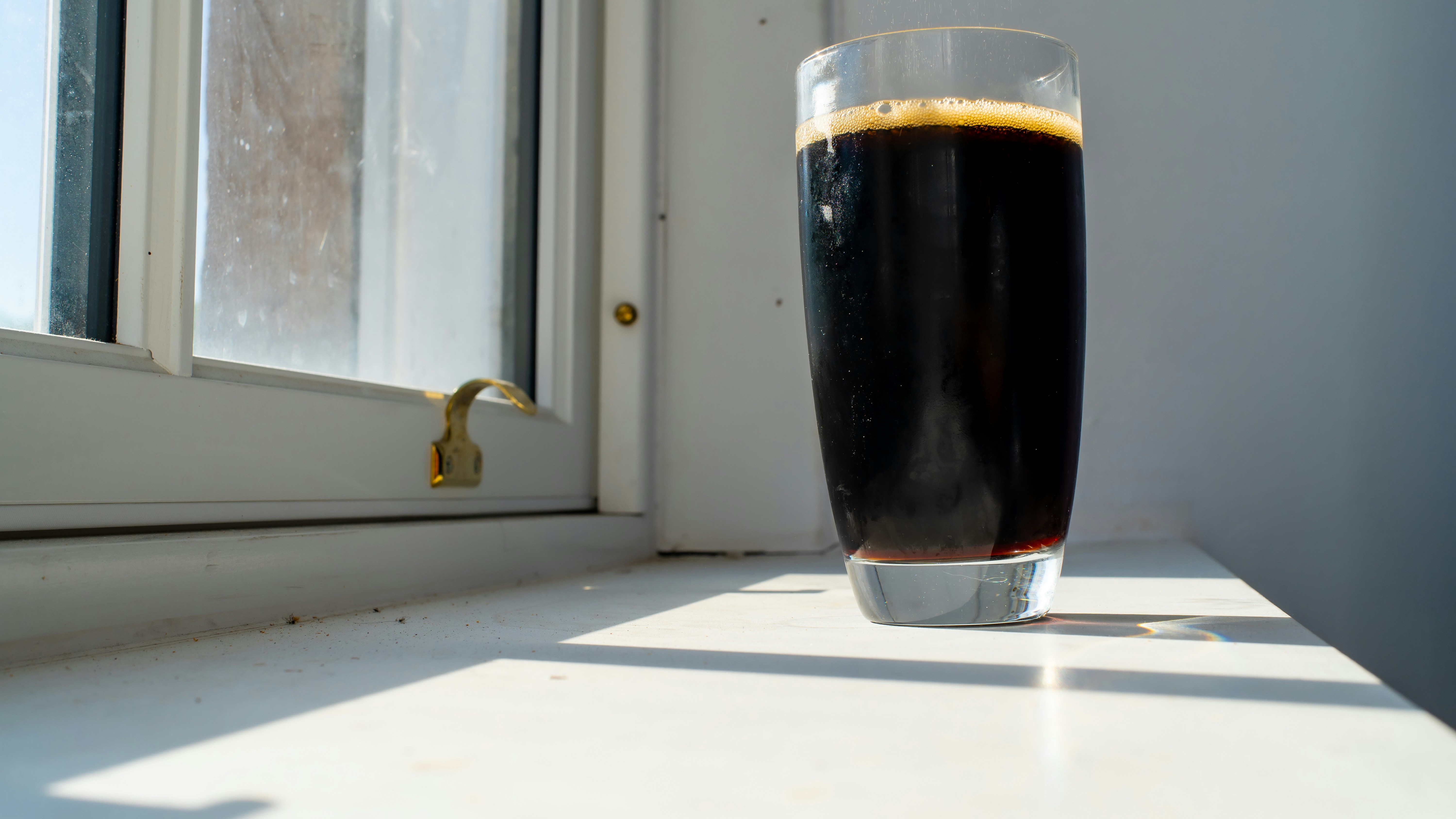 A glass of dark beer sits on a sunlit windowsill.