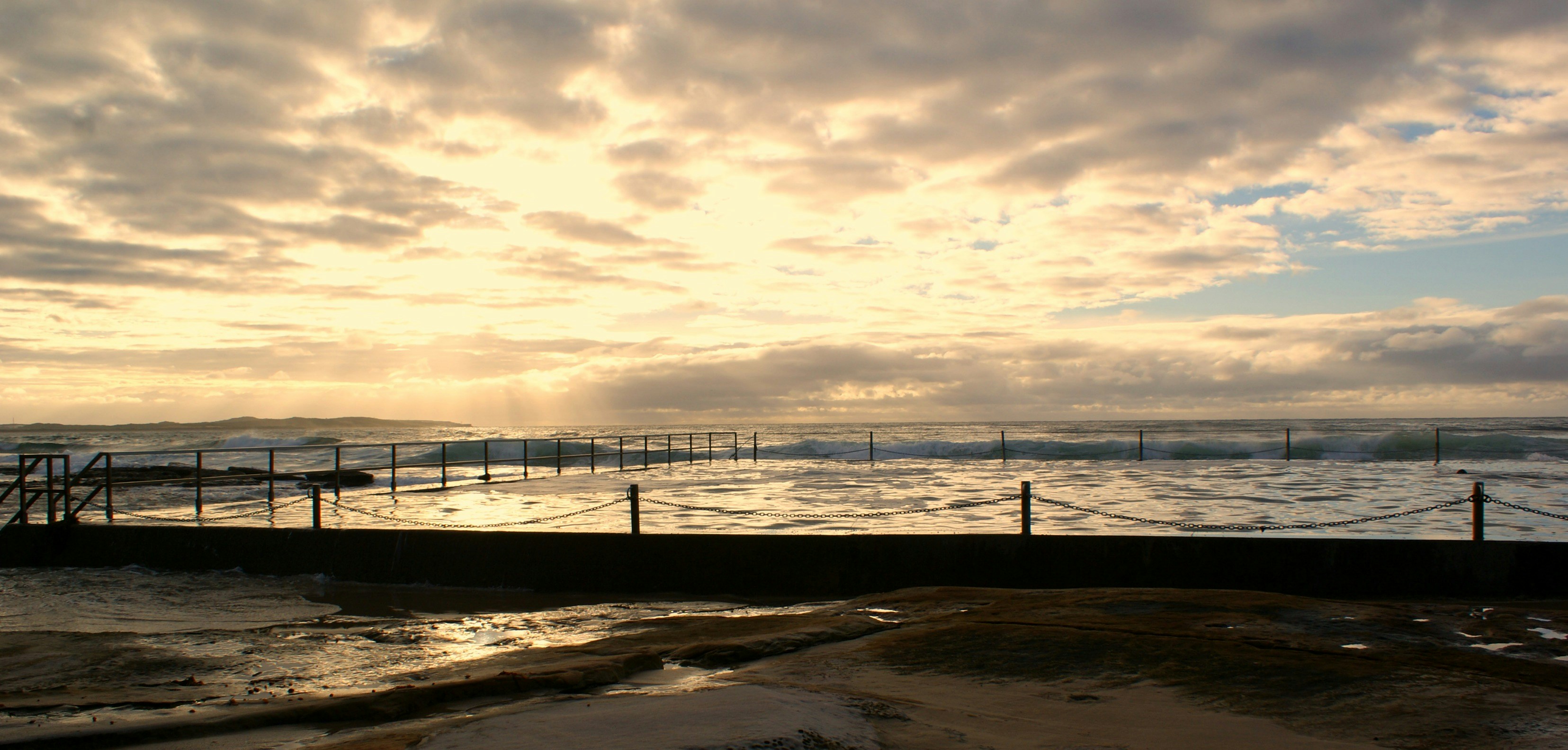 Golden light over ocean waves and a sea pool wall.