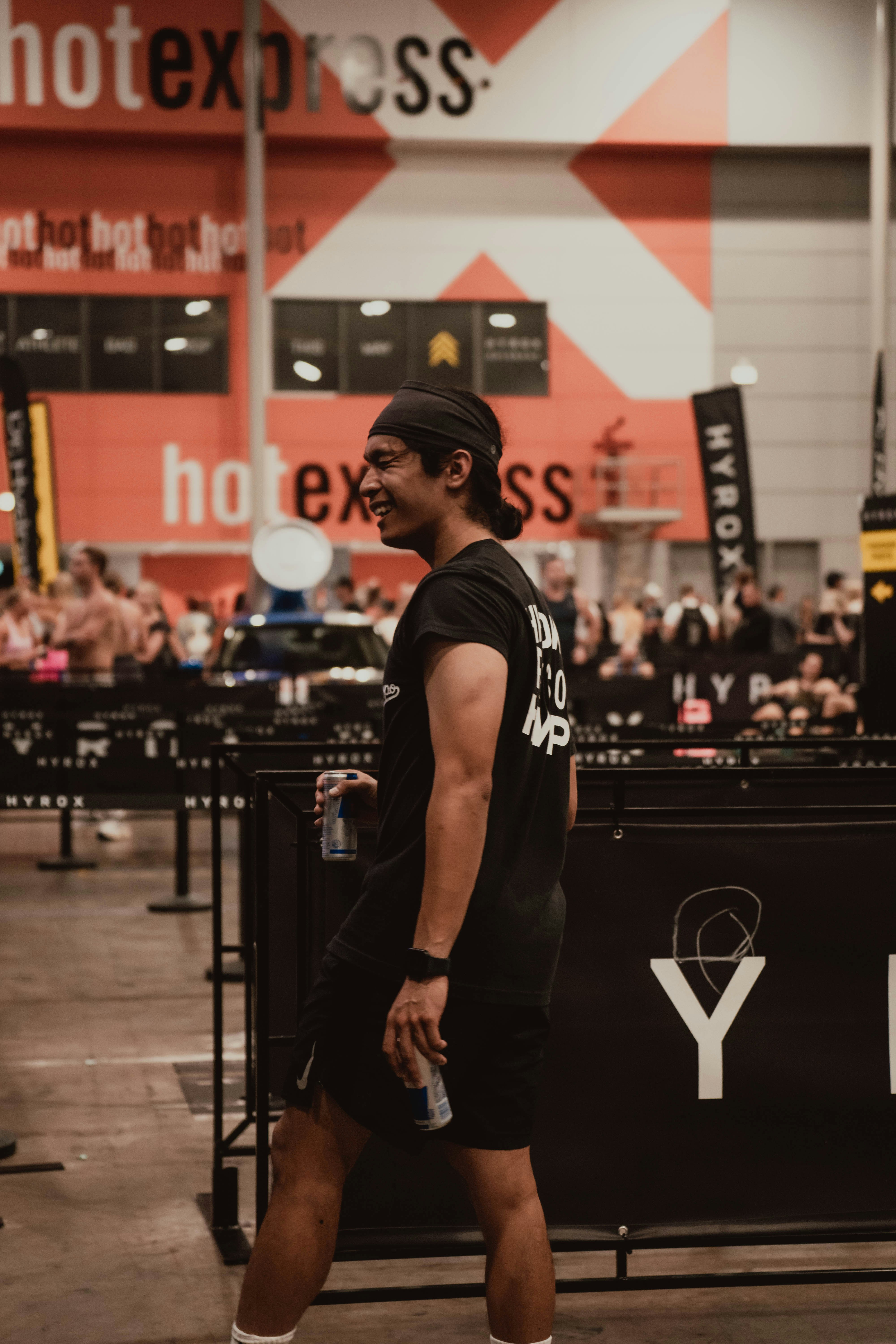 Side-profile at the Expo: a wristbanded athlete holds a drink amid a sea of banners