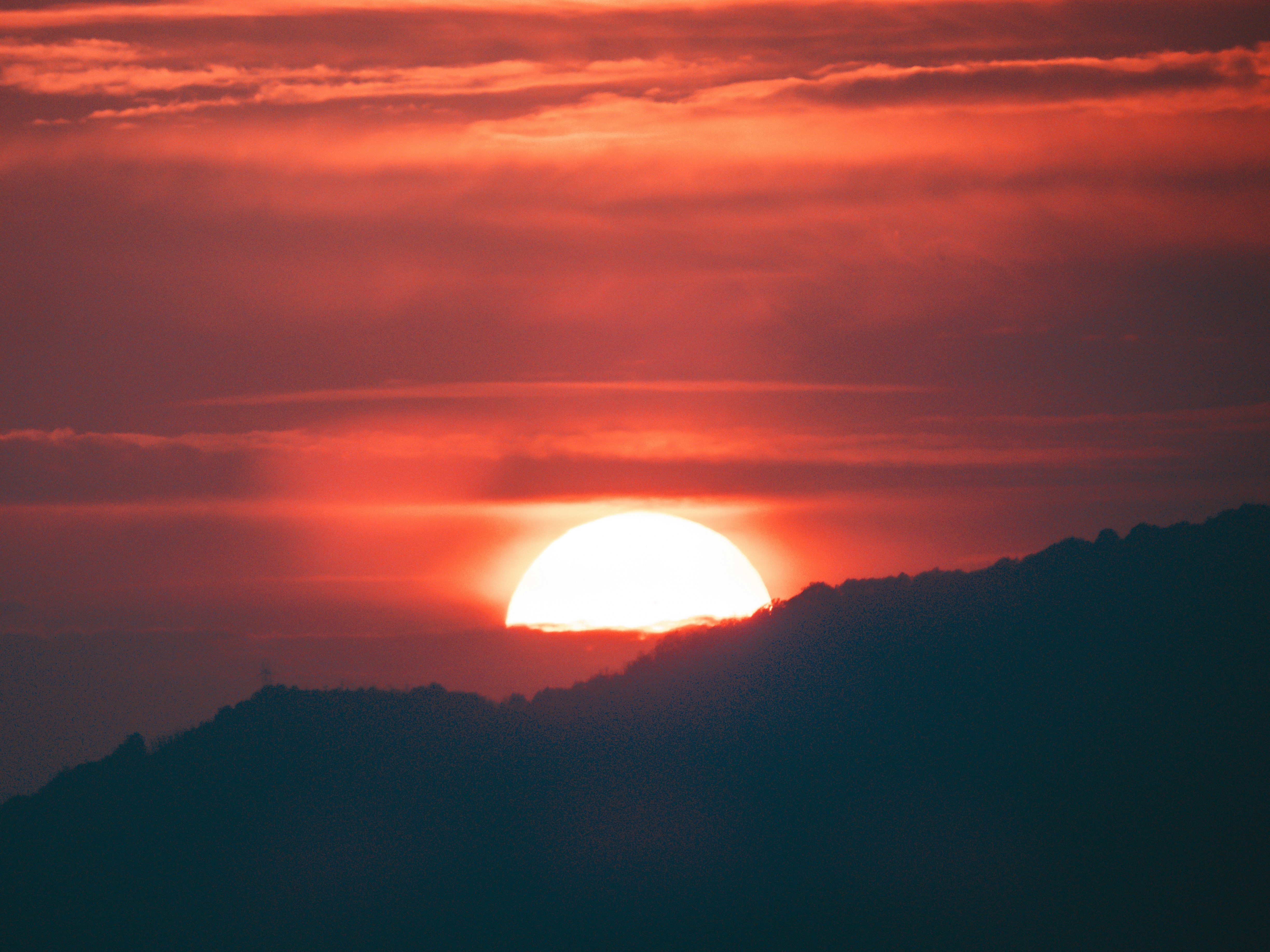 Dramatic crimson sunset over a dark mountain silhouette. The sun dips below the horizon, painting the sky and layered clouds in deep shades of red and orange, framed by dark foreground foliage. Perfect for intense, powerful backgrounds and moody aesthetic covers.