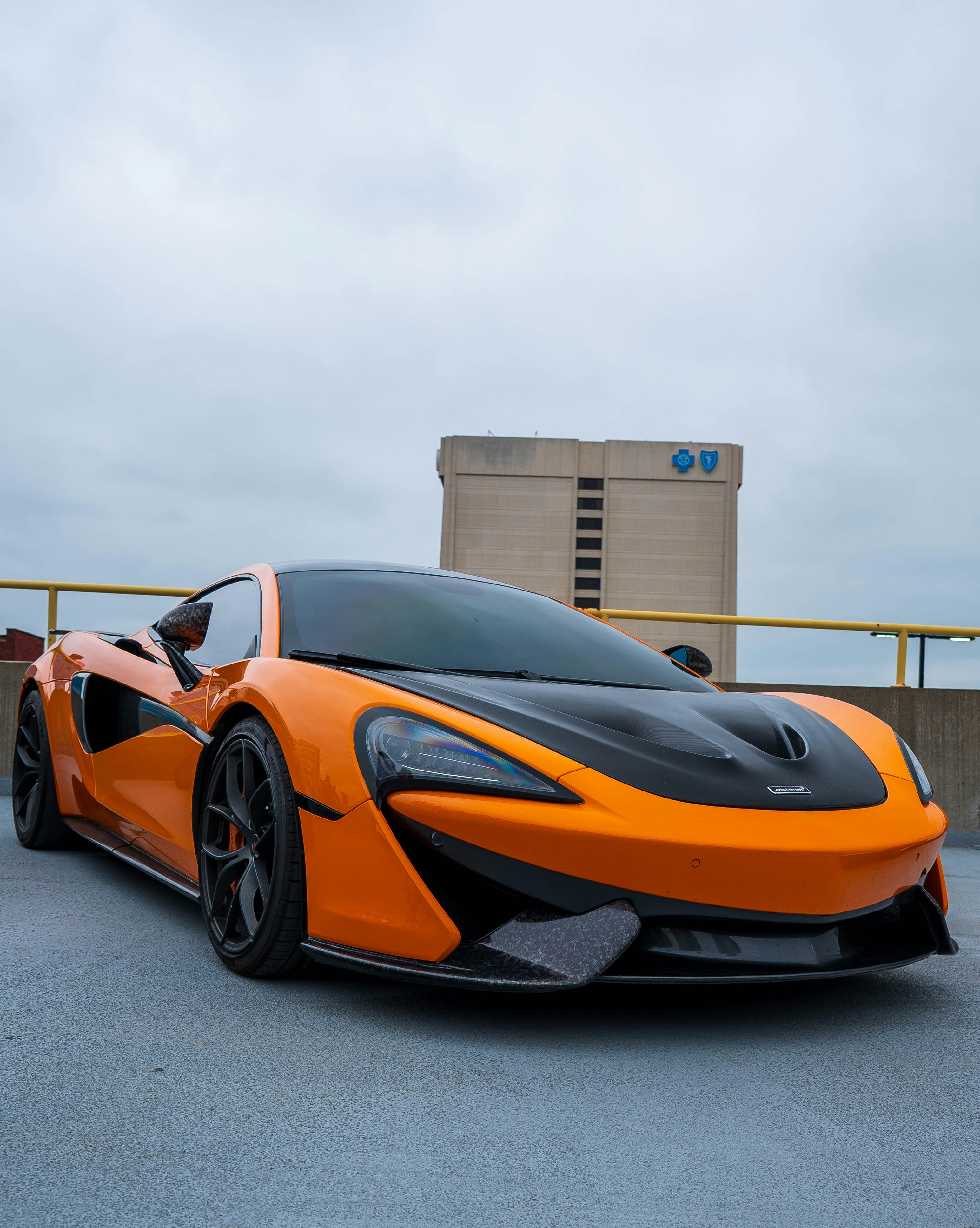 Orange sports car parked on a rooftop with a cloudy sky and industrial backdrop, emphasizing lines and stance.