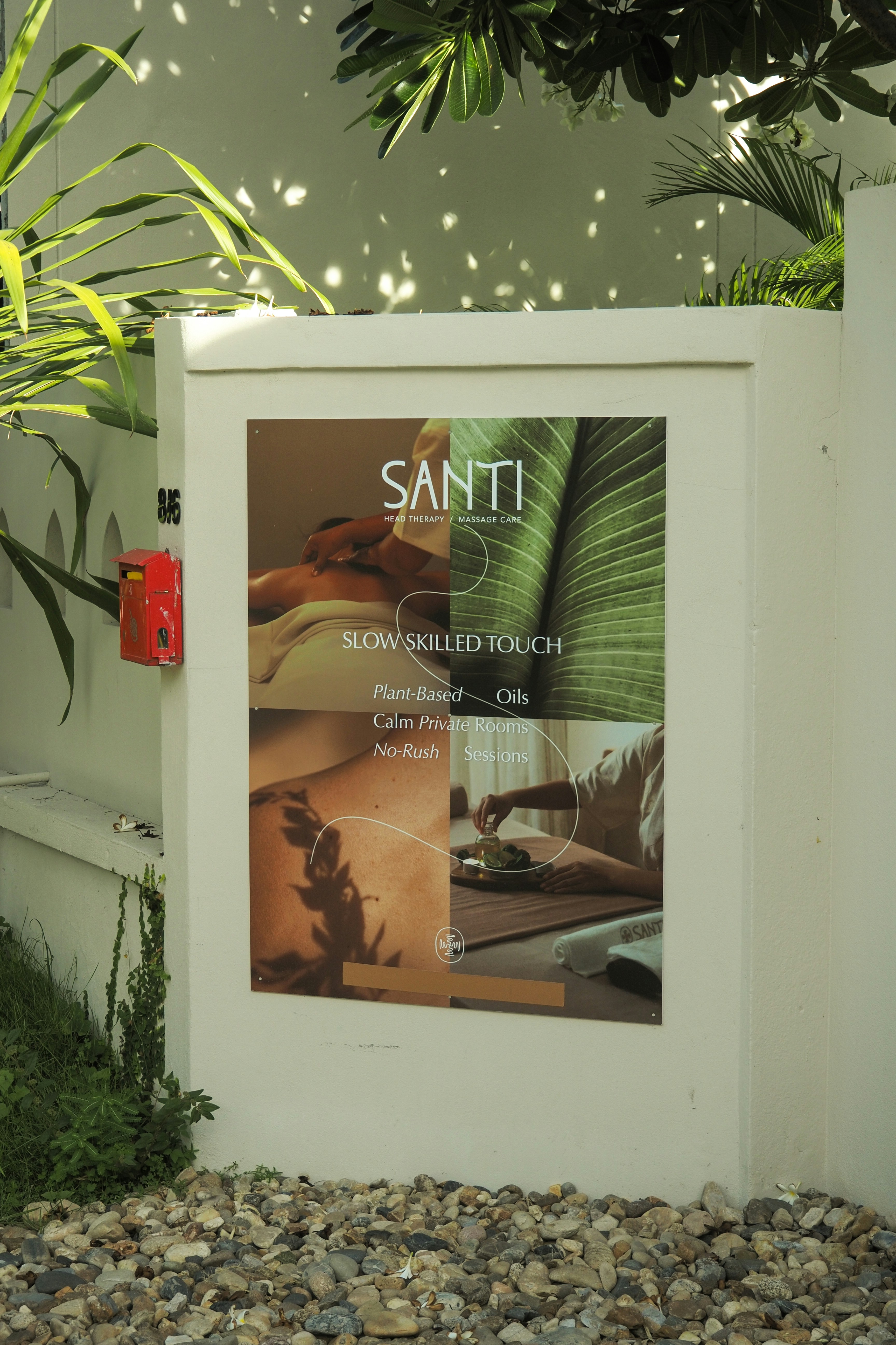 Wellness poster mounted on a white wall framed by tropical greenery and pebbled ground; a calming spa ad with garden setting.