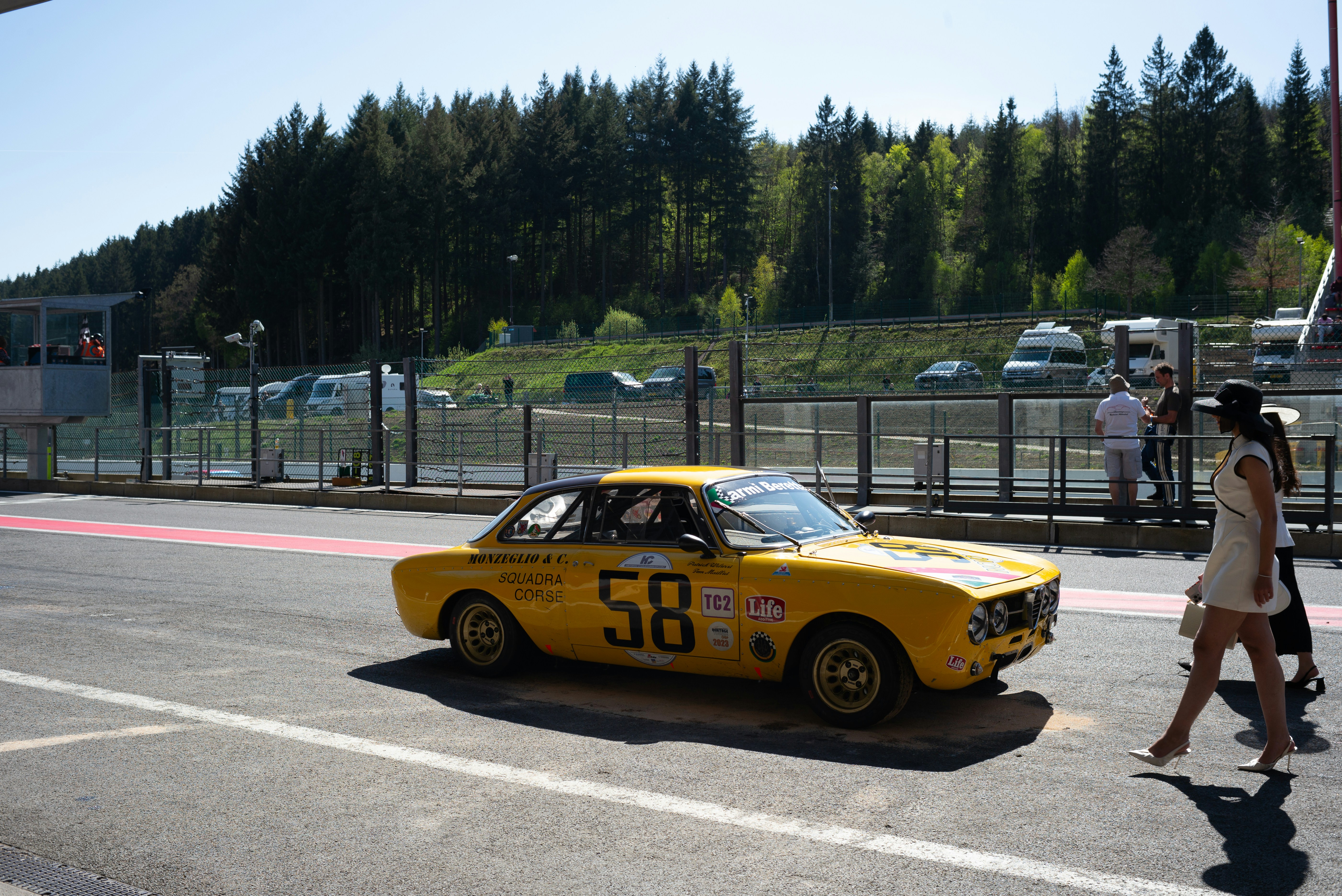 Yellow race car crossing the sunlit track as spectators walk along the pit side