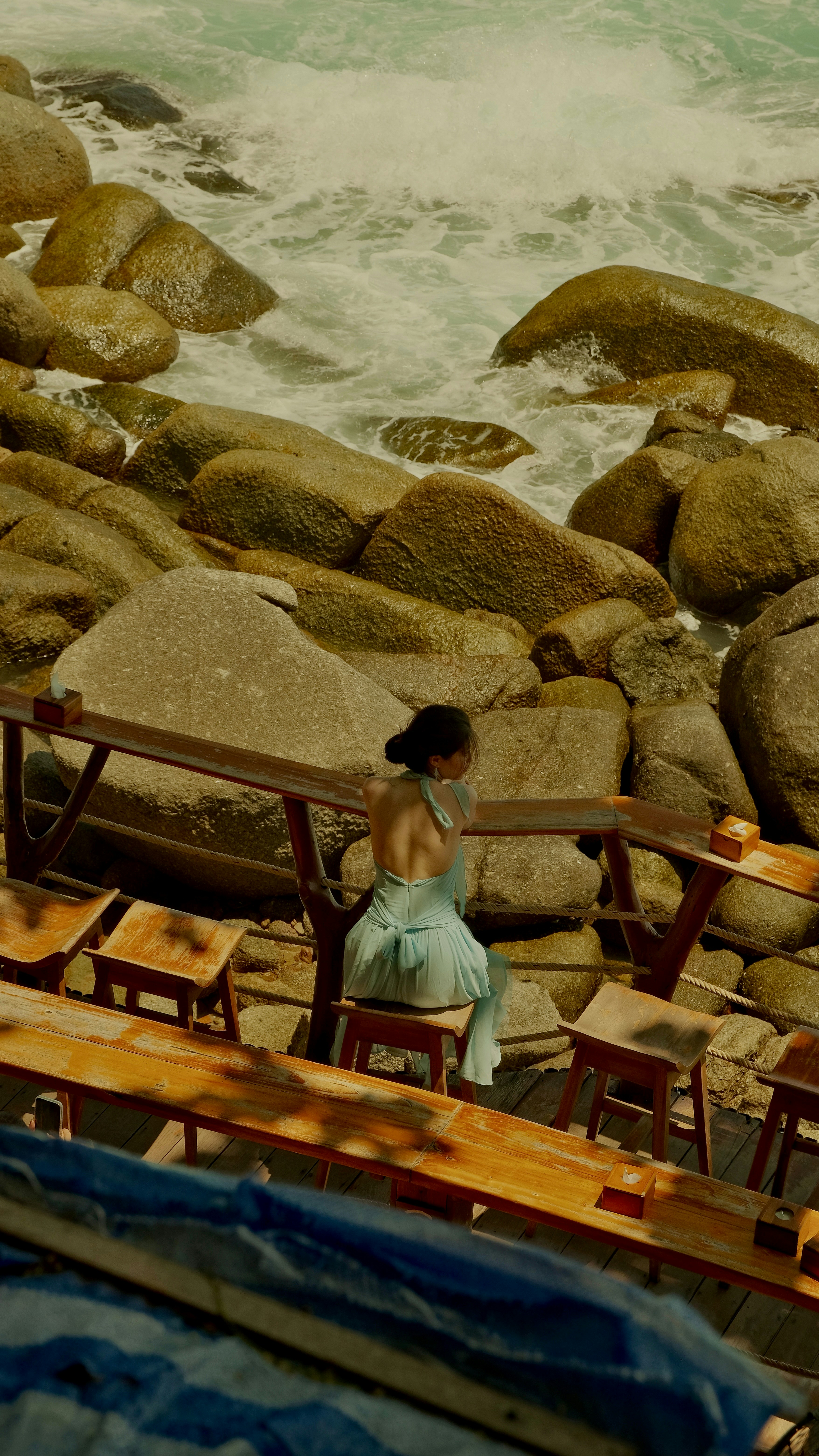Coastal Terrace: A lone dancer in pale turquoise overlooks sea-strewn rocks from sunlit benches