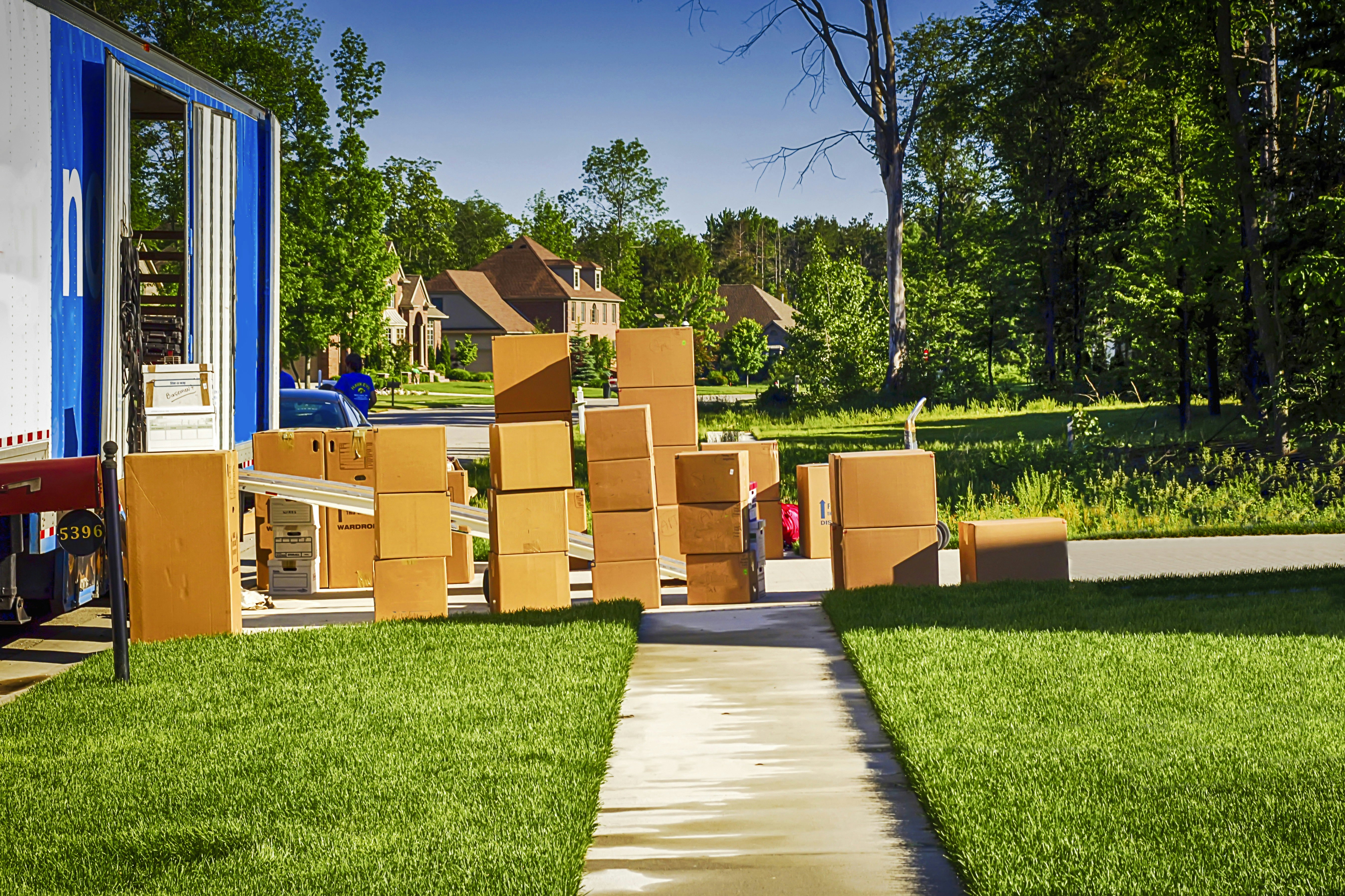 A row of stacked cardboard boxes sits along a concrete path beside a blue truck in a sunlit residential yard, with green lawns and trees in the background.