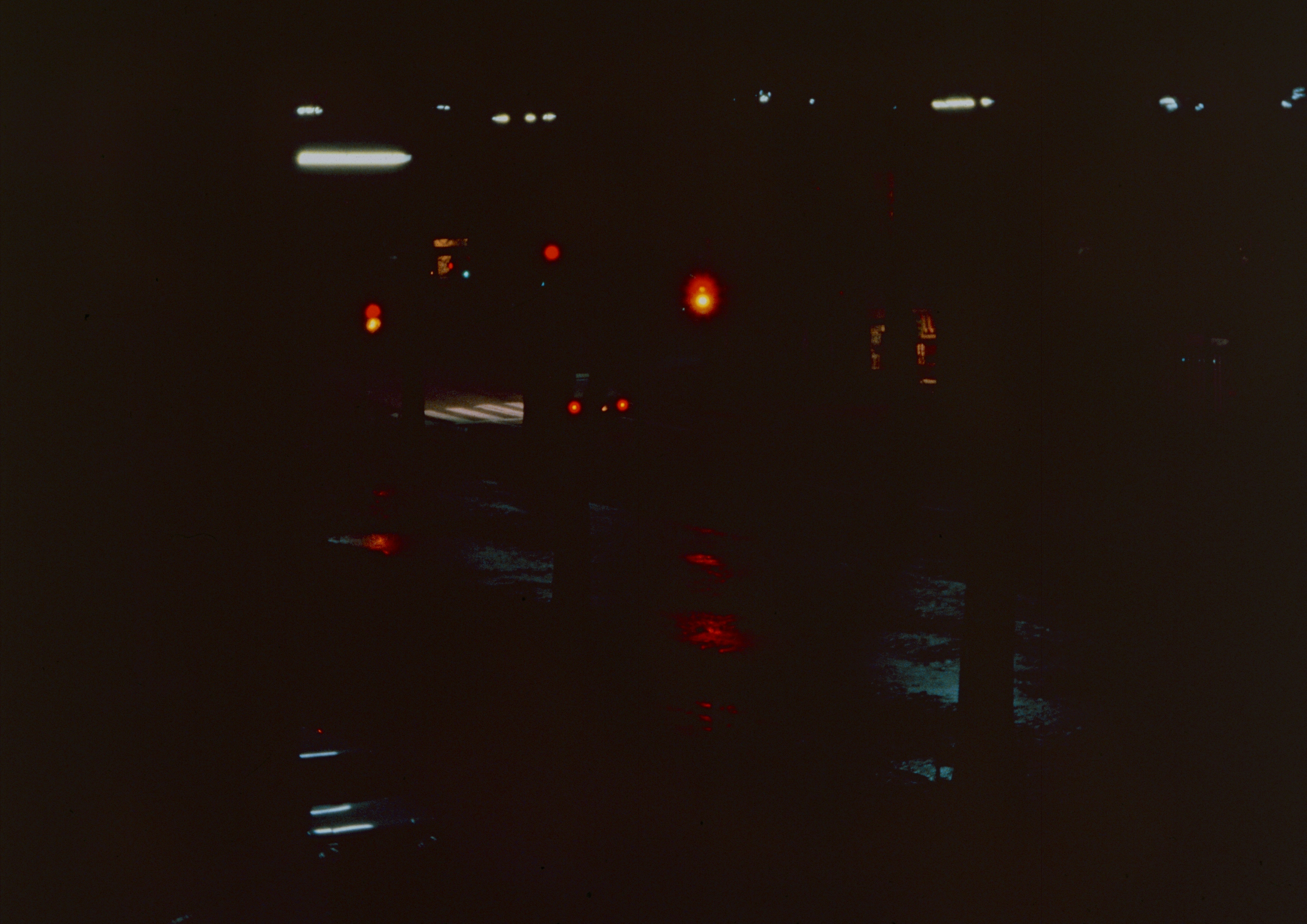A darkened night scene at the Sønder Boulevard and Dybbølsgade intersection glows with scattered points of color—amber traffic lights, red brake lights, and cool blue-green reflections shimmering on the rain-soaked street. A faint white streak of a passing headlight cuts through the lower frame, while a crosswalk's pale stripes emerge faintly in the gloom. The heavy shadows and diffused glow give the image an almost abstract, moody quality. It's a quiet, atmospheric glimpse of a rainy Vesterbro evening in late-1970s Copenhagen, long after the streets have emptied.