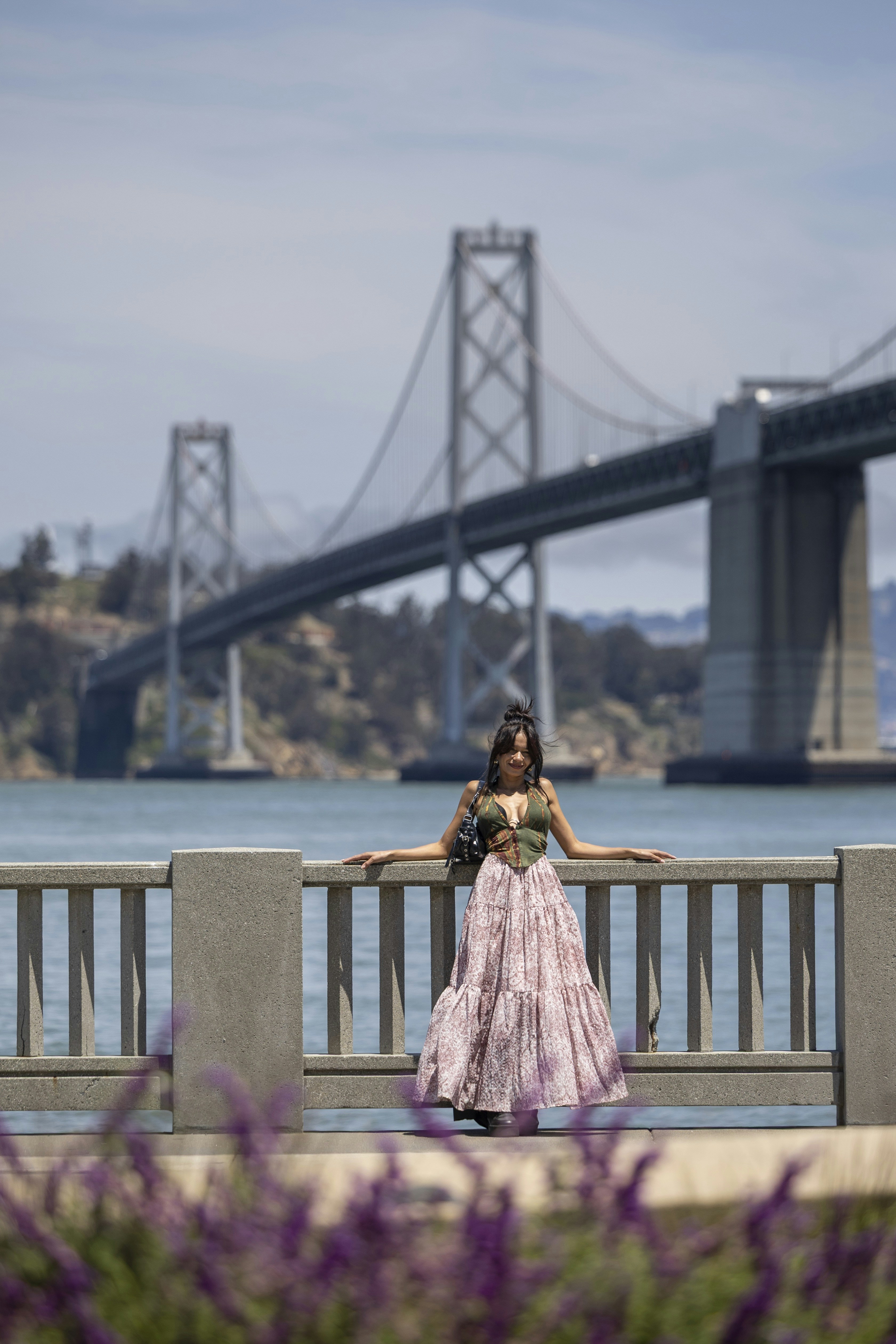 Elegant portrait of a woman in a flowing pink dress standing at a railing with a large suspension bridge in the background, water beyond, and distant coastline visible. Photograph type: candid fashion portrait.