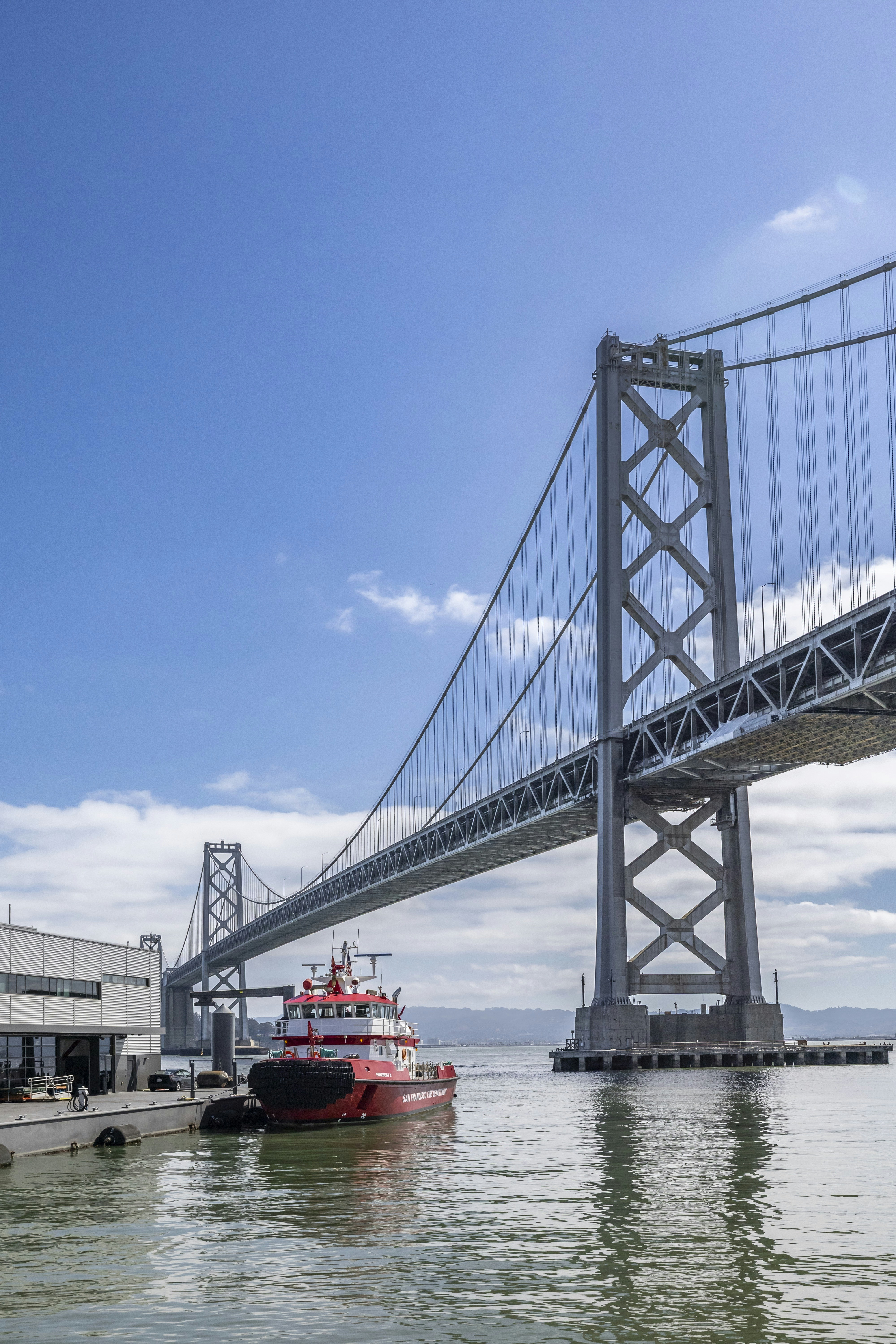 A red tugboat docked under a large suspension bridge.