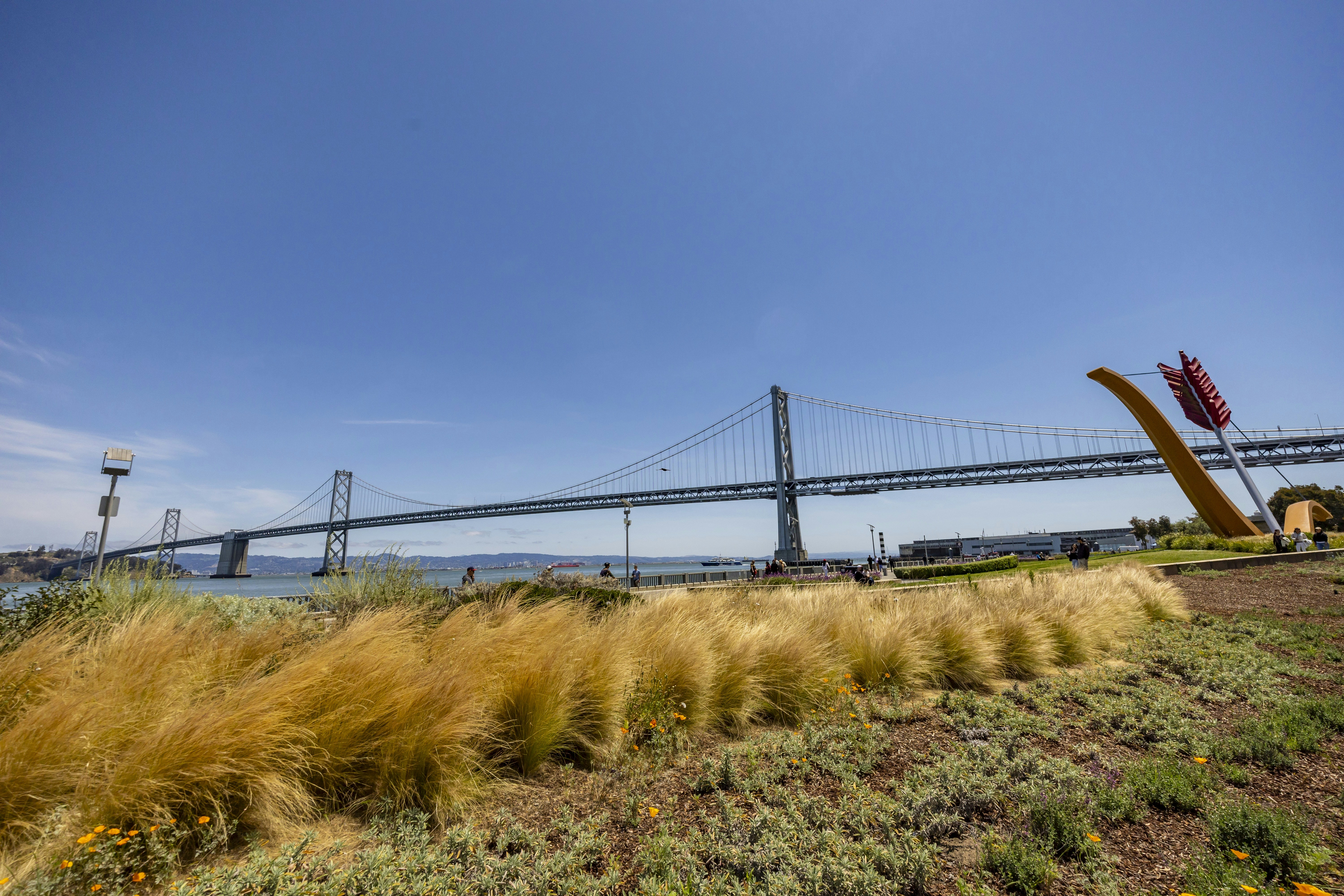 Bay bridge, golden grasses, and sculpture under a clear sky.