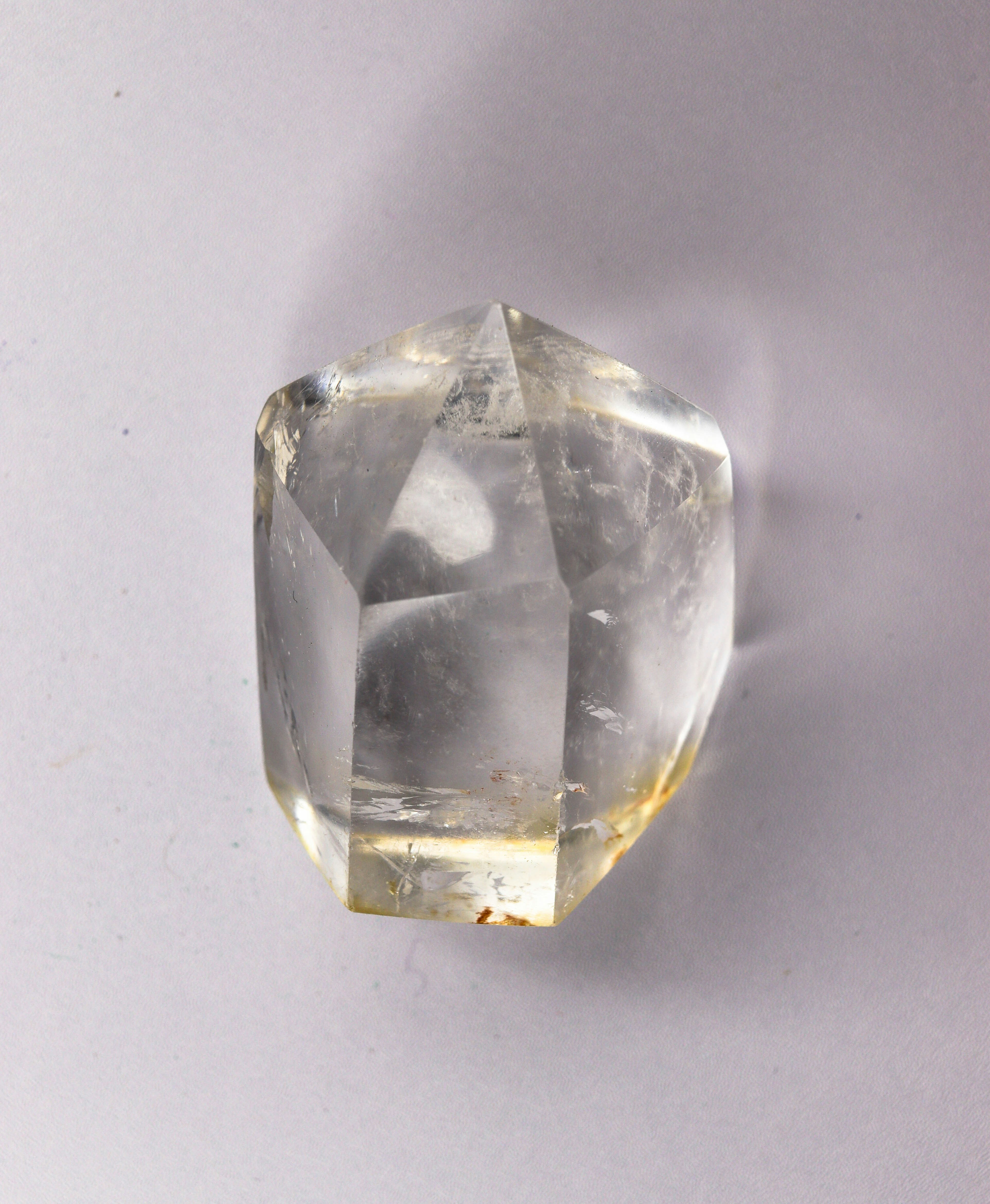 Clear crystal gemstone sits on a light surface, its multifaceted faces catching soft light and revealing subtle color hints. A study in geometry and refraction.
