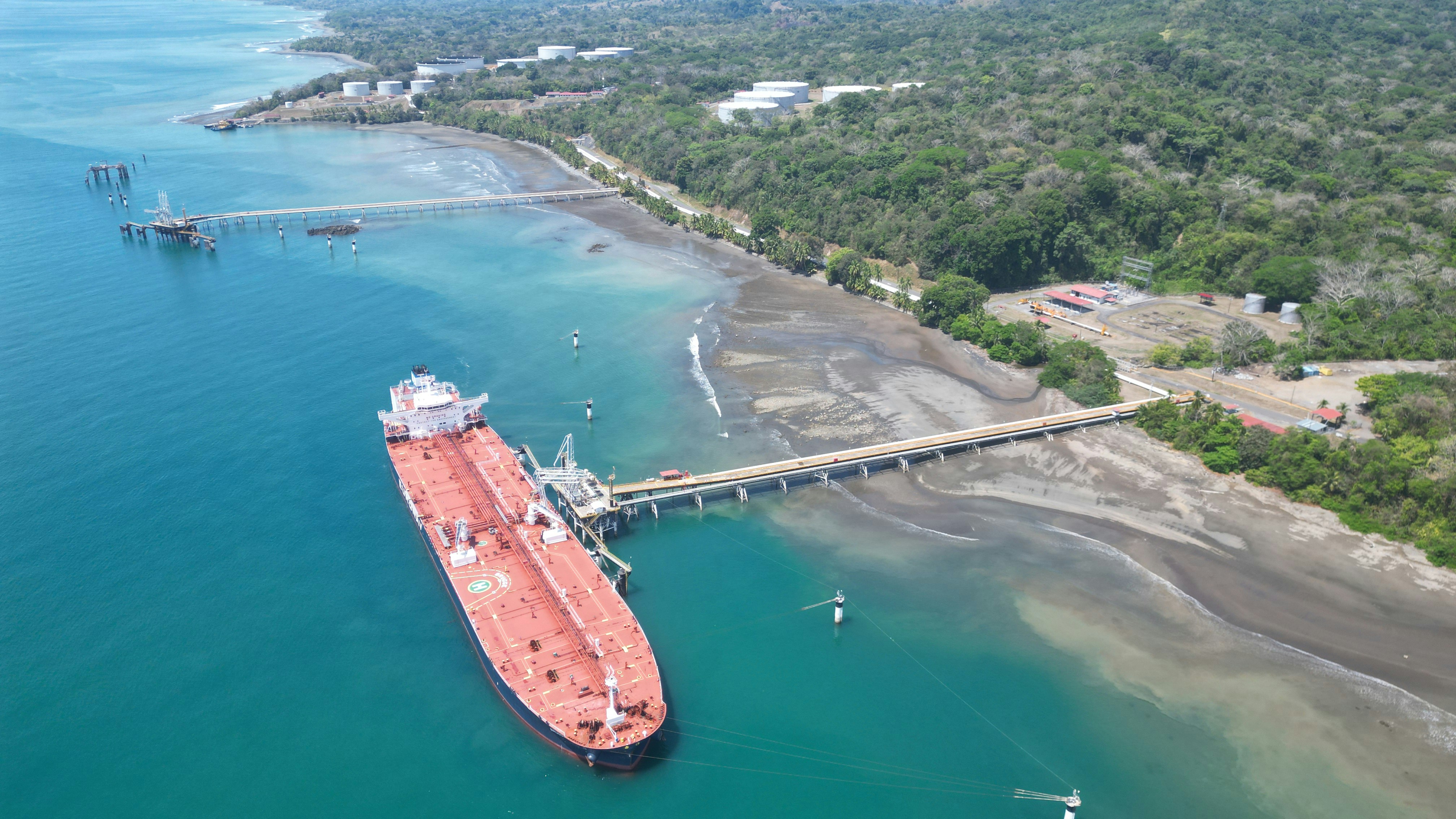 Red-hulled tanker at a coastal pier, caught mid-drift against turquoise shallows